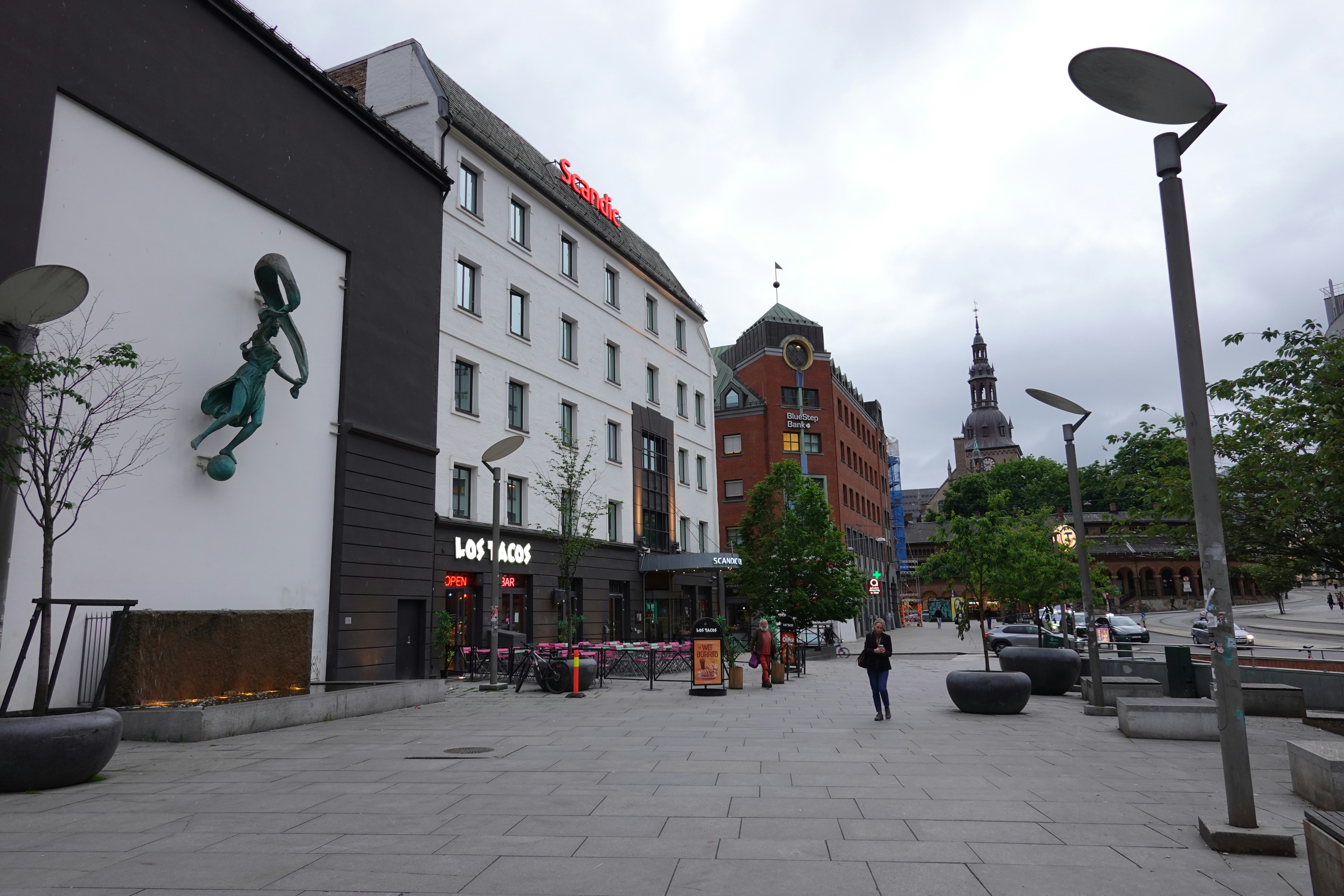 Wide pedestrian street with a bronze sculpture on the left, modern storefronts, and a distant church tower under a cloudy sky.