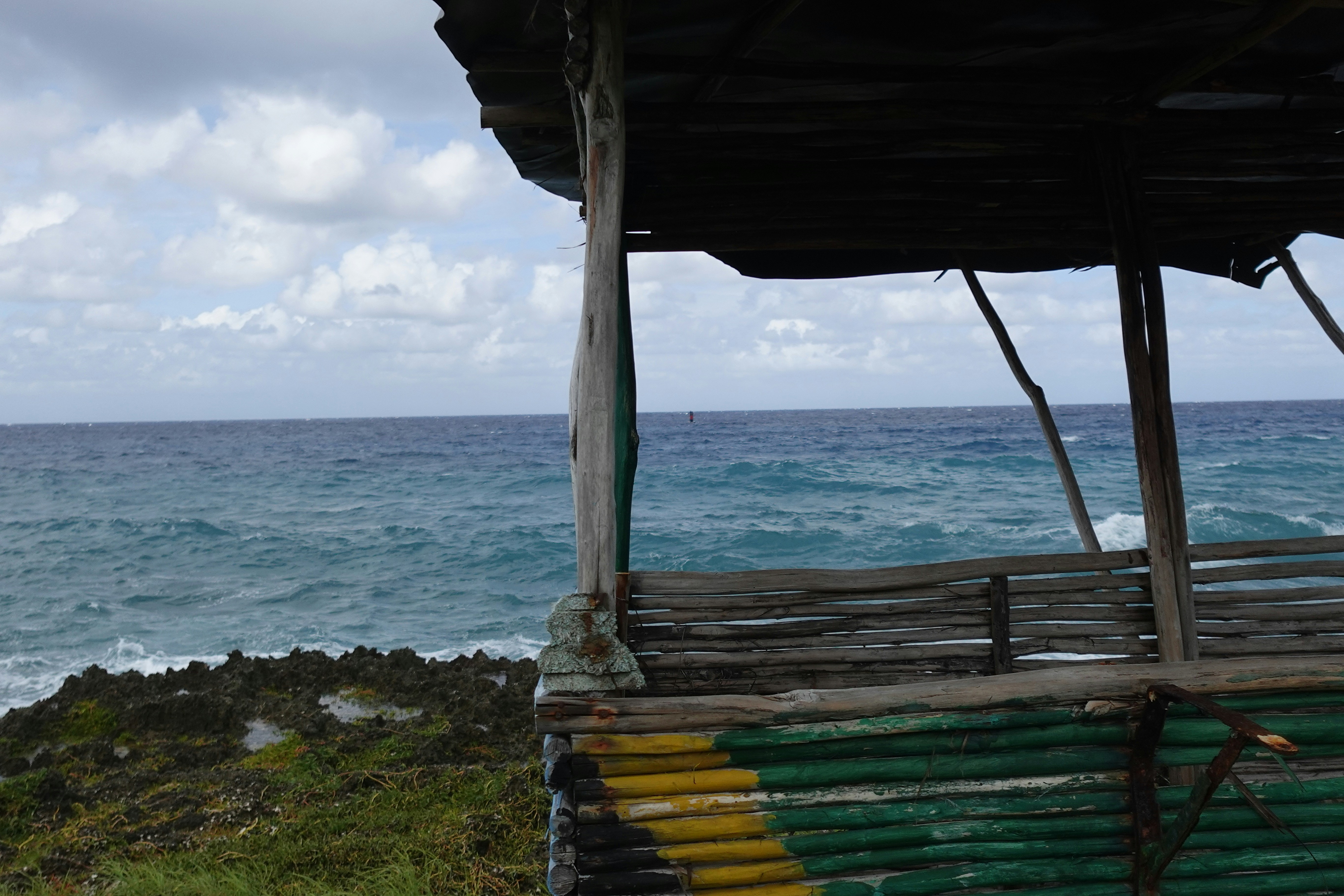 Shade pavilion by the rocky shore with a colorful wooden bench facing the sea, under a broad sky and wispy clouds.