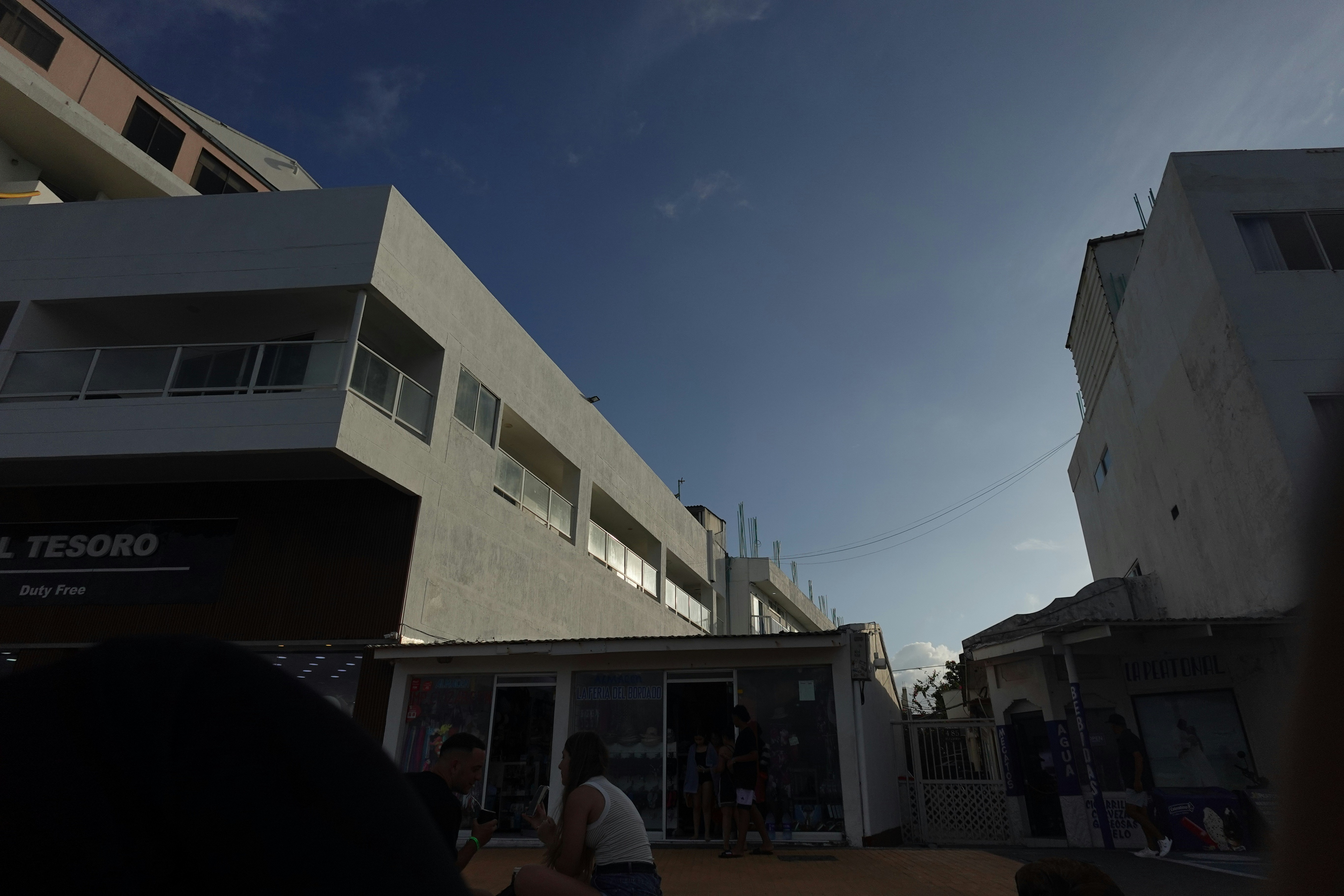 Wide-angle street scene with modern buildings framing a shaded plaza and a few people gathered near the storefronts; the sky dominates the upper portion of the frame.