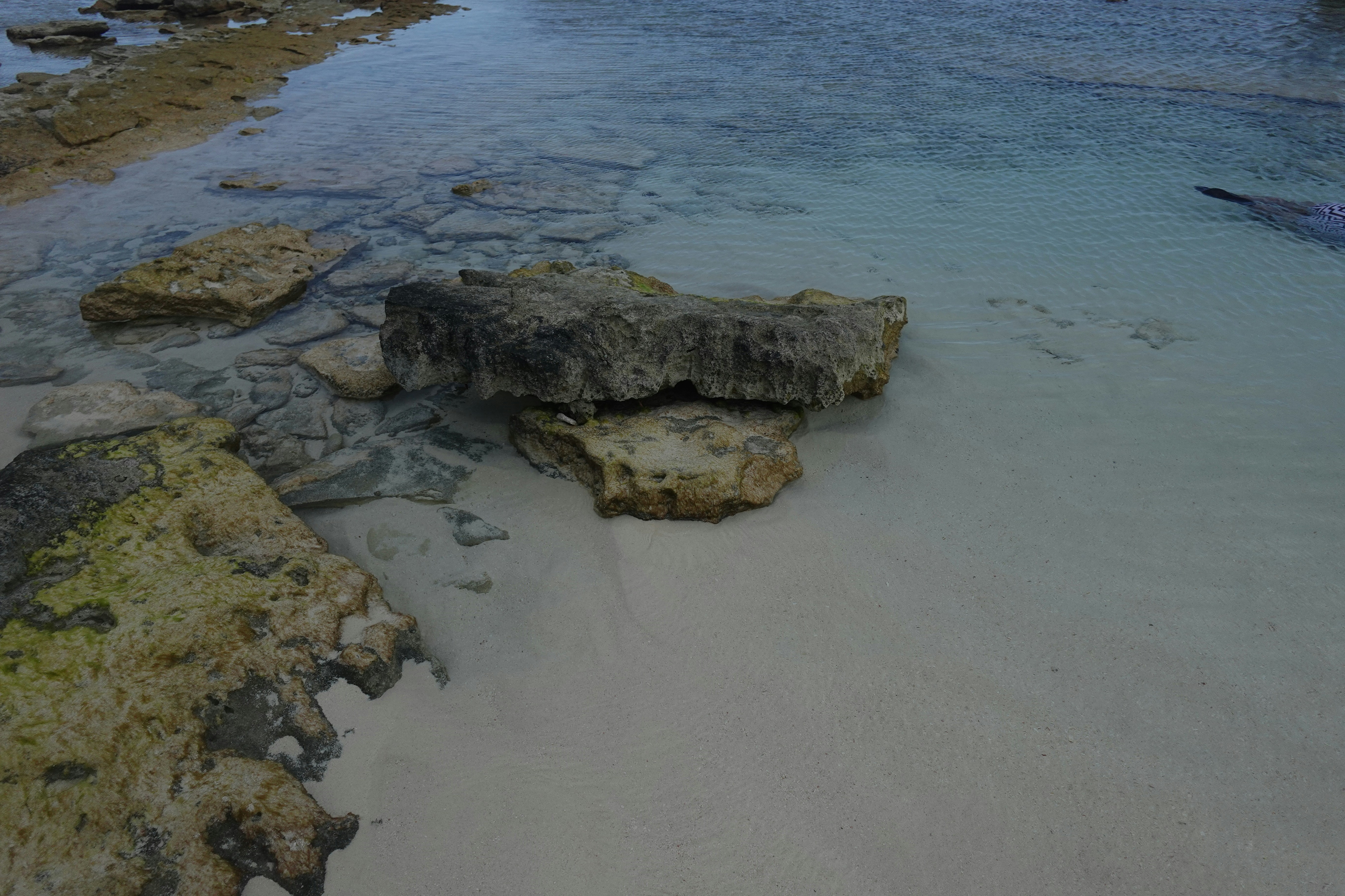 Soft beach with layered rocks meeting calm turquoise water along a pale sand shore. A tranquil coastal scene with subtle color gradation and gentle waves.