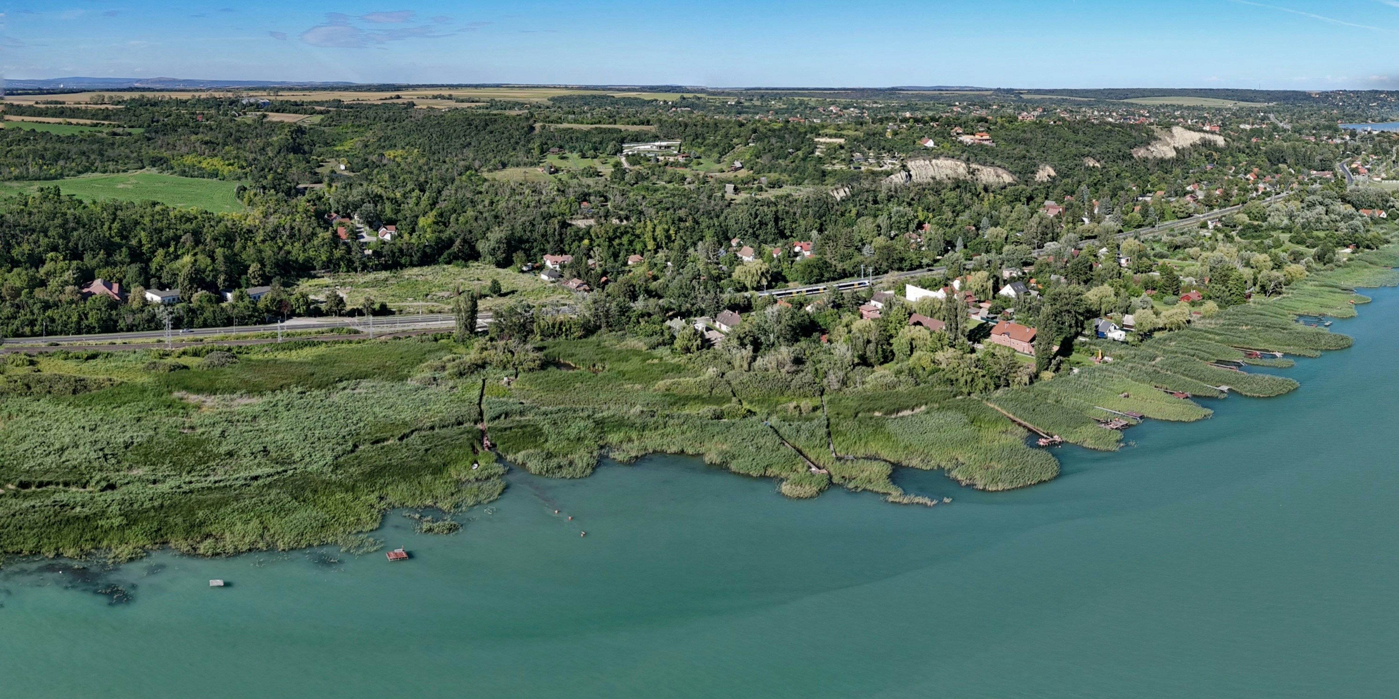 Aerial view of the northern shore of Lake Balaton, Hungary, featuring blue waters, lush green wetlands, reed beds, and a peaceful countryside landscape under a clear summer sky.