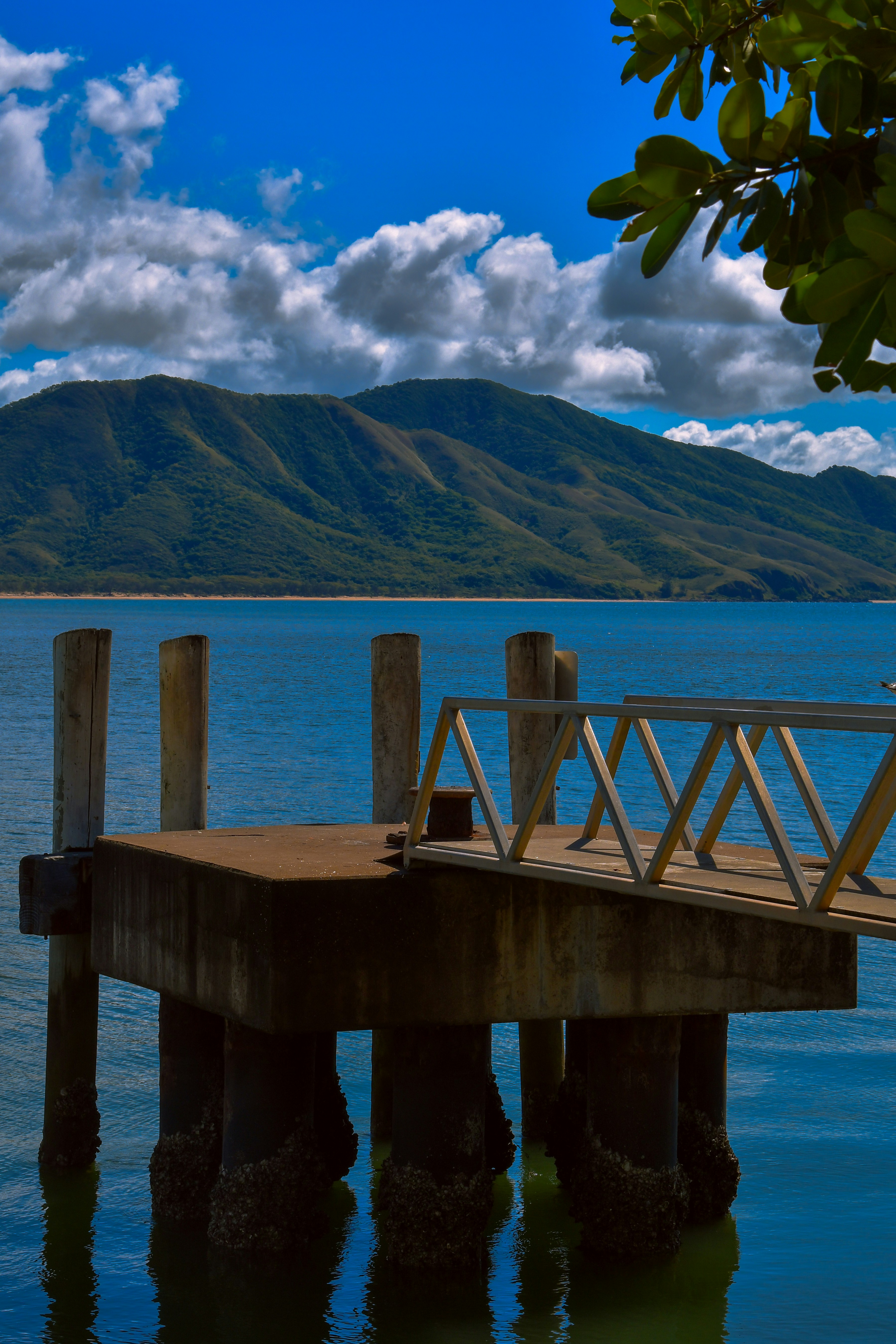 Concrete pier over water with mountains and blue sky.