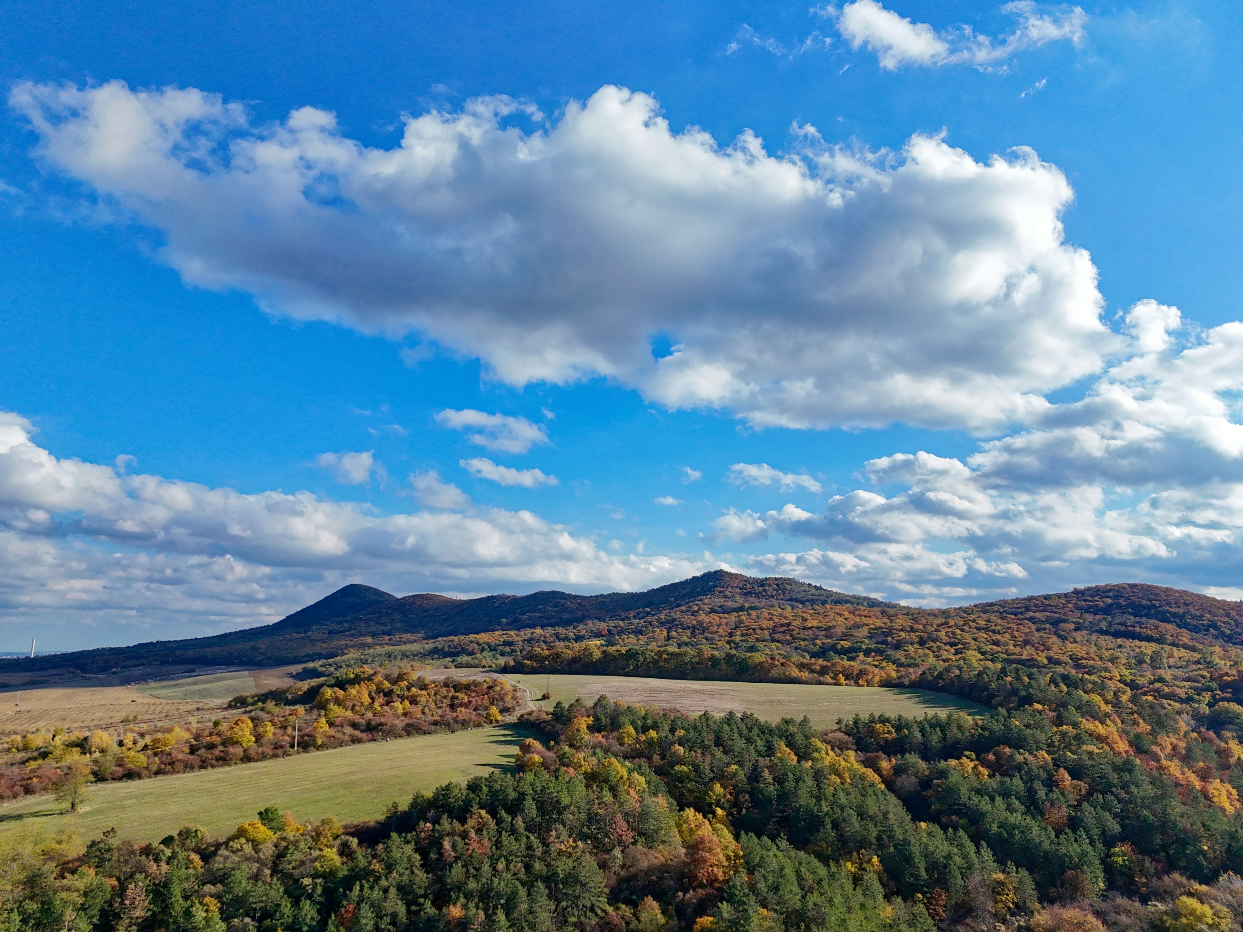 Scenic aerial view of autumn hills and a vibrant blue sky with fluffy clouds in Bicske, Hungary. The landscape shows a mix of green pine trees and colorful fall foliage covering the rolling hills under a bright, sunny sky.