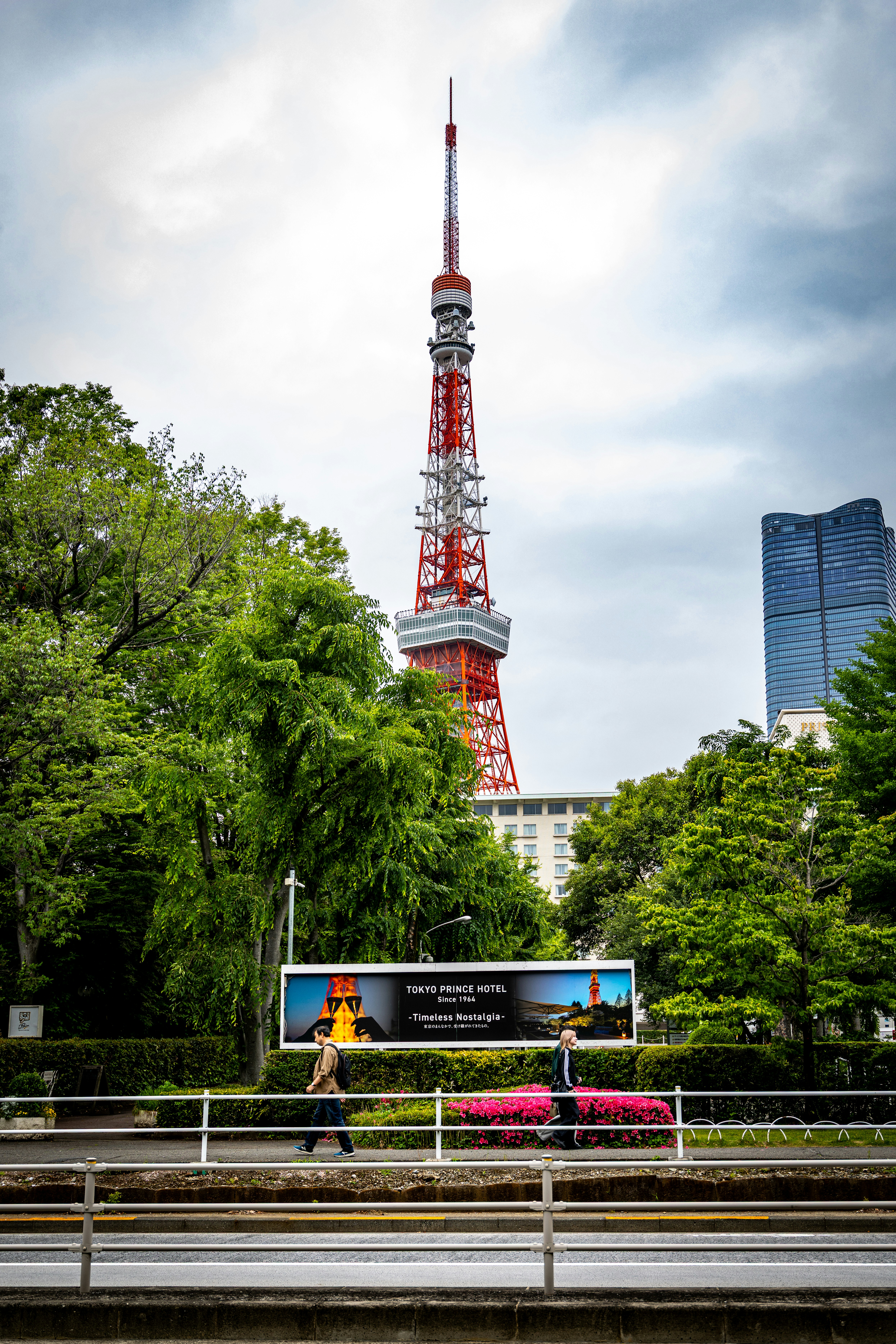 Towering Tokyo Tower rises above lush trees and a cityscape, framed by a quiet street and a billboard in the foreground.