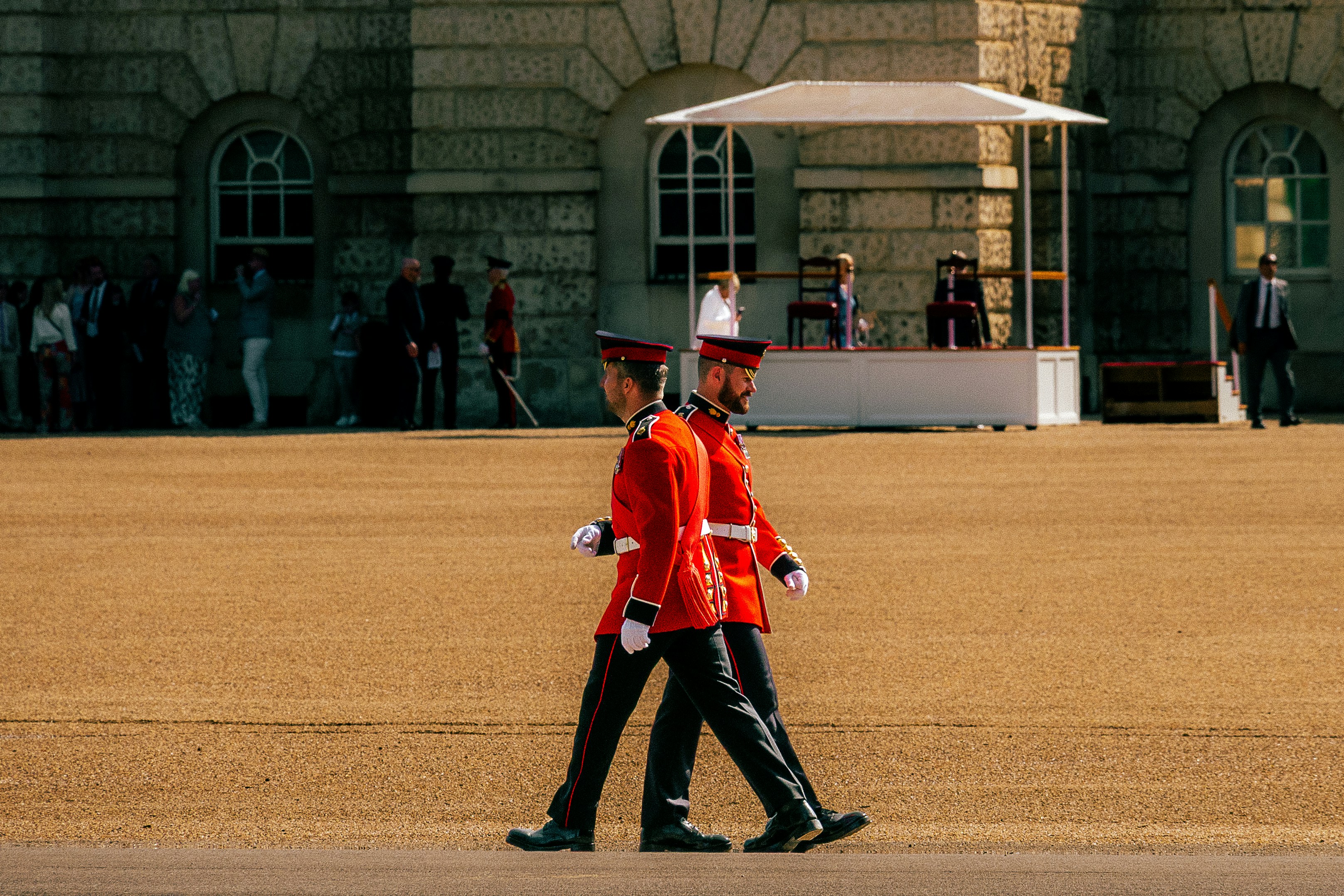 Two soldiers in red uniforms march past a building.