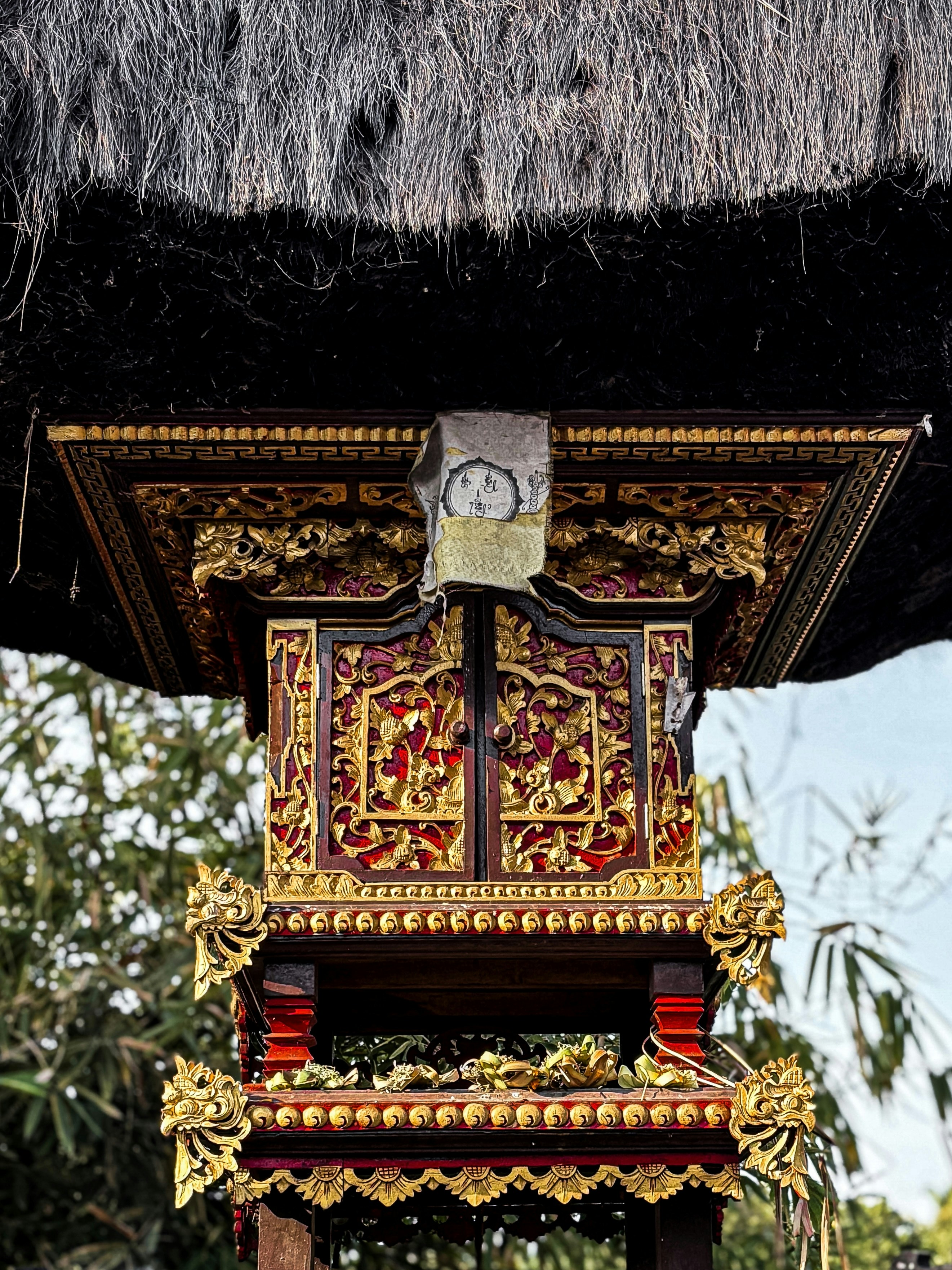 Intricately gilded paneling atop a carved pedestal, framed by hanging thatch, with lush greenery in the background.