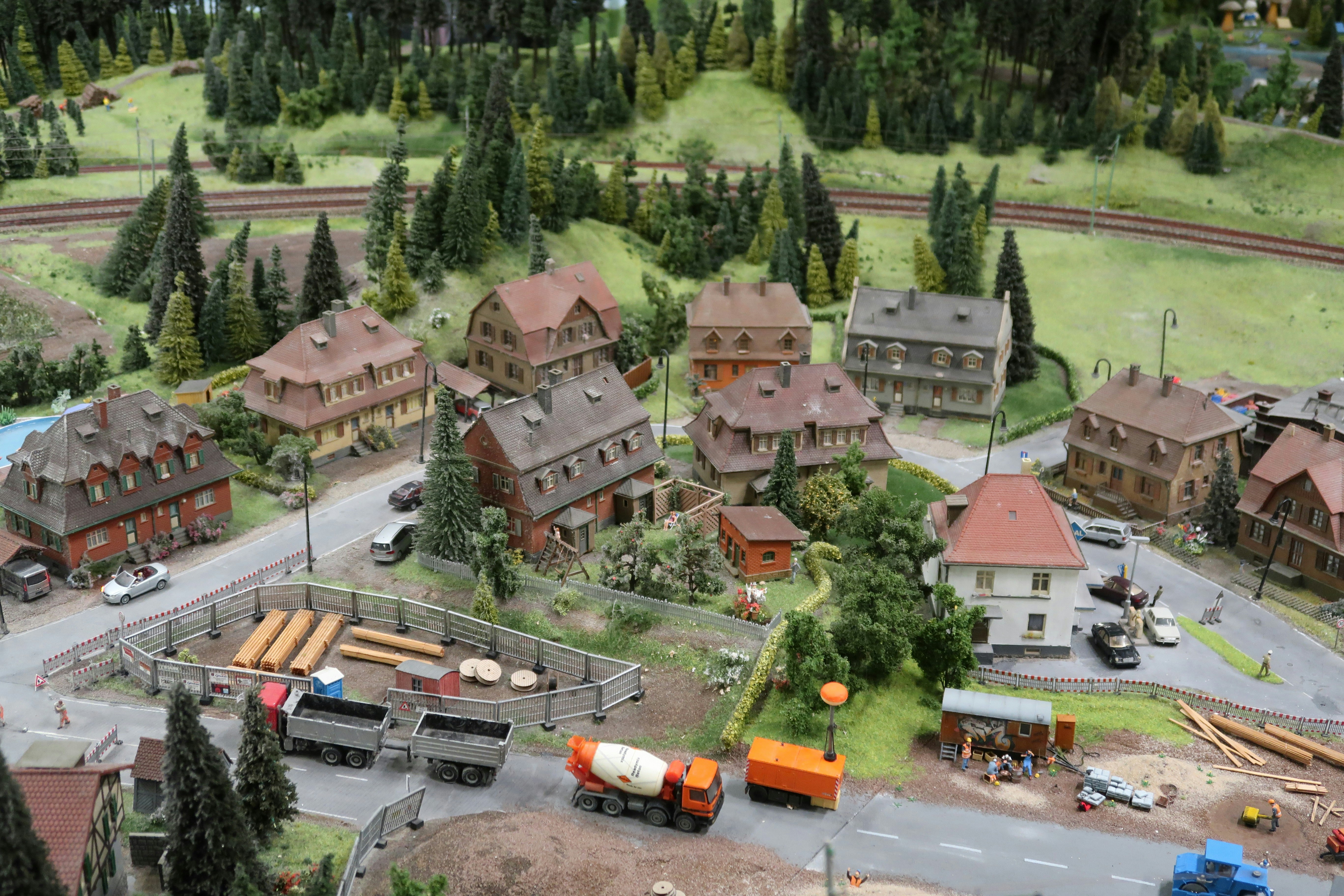 A detailed model village with clay-toned houses, trees, and a small industrial yard featuring trucks and equipment, captured from an elevated angle. The scene emphasizes scaled residential life and rail-adjacent activity.