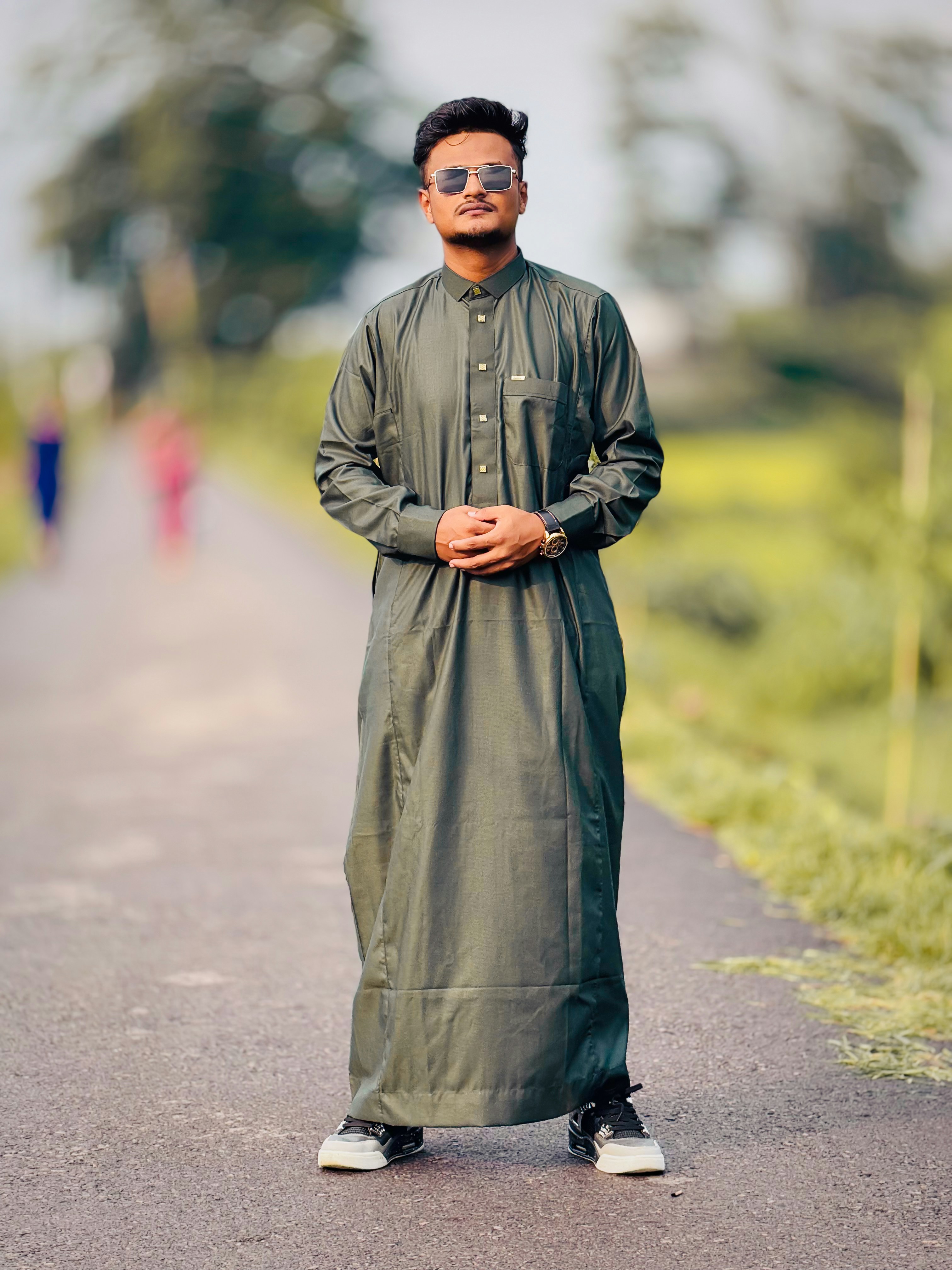 Man in olive thobe stands centered on a park path, hands clasped, with a blurred green landscape in the background. The setup emphasizes a clean full-body portrait.