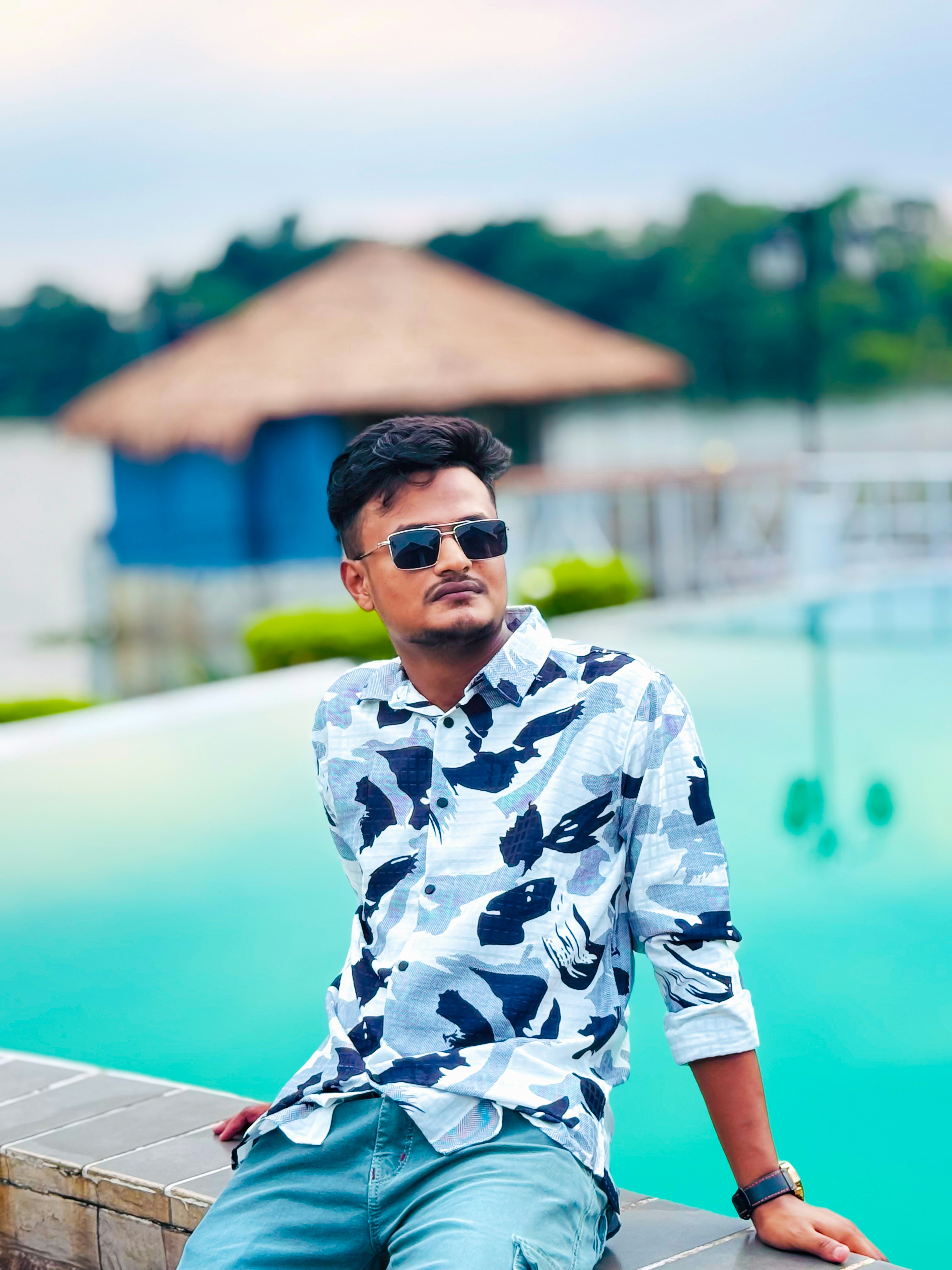 A man in sunglasses sits by a swimming pool.