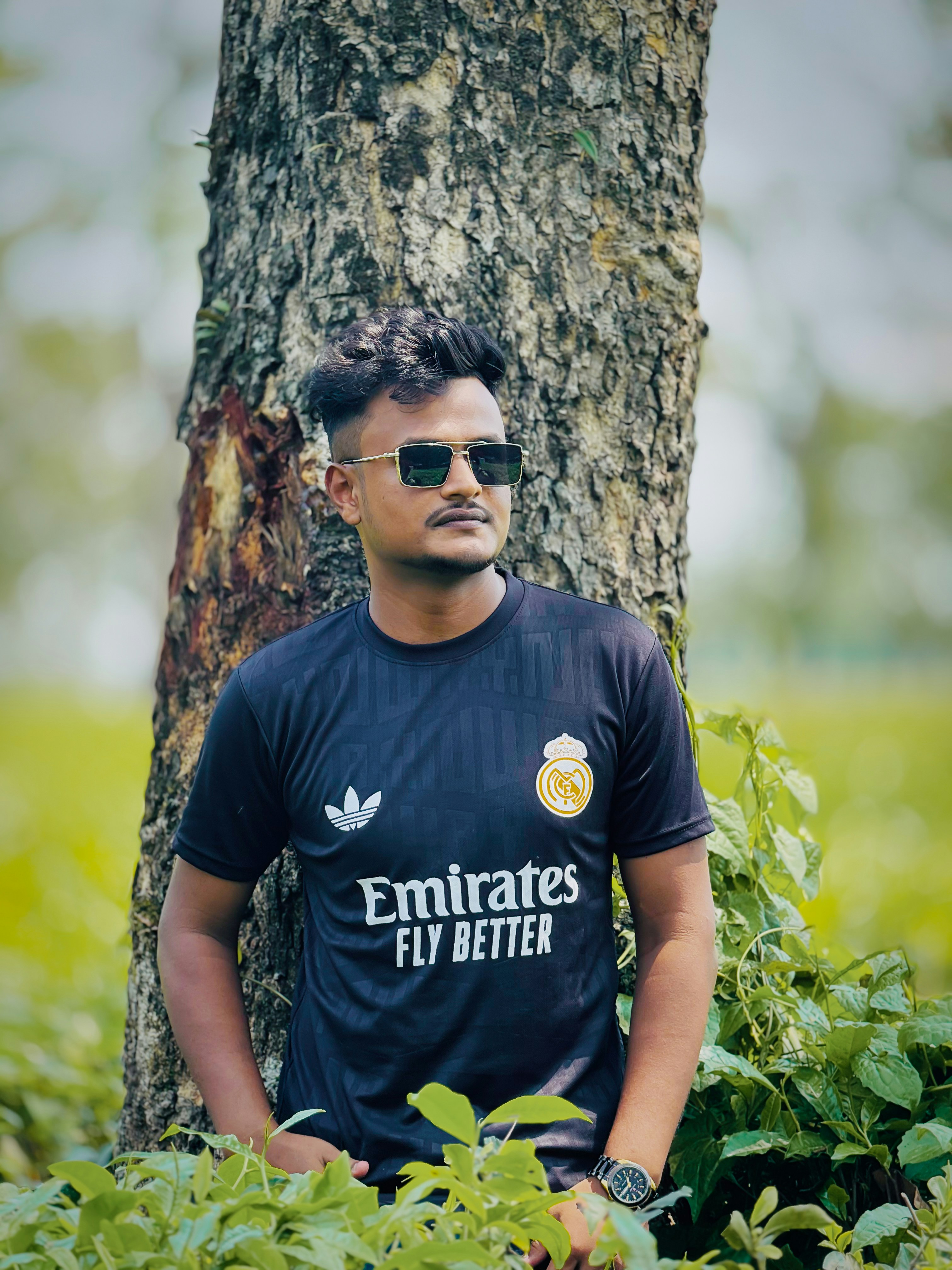 Young man in sunglasses leans against a tree outdoors.