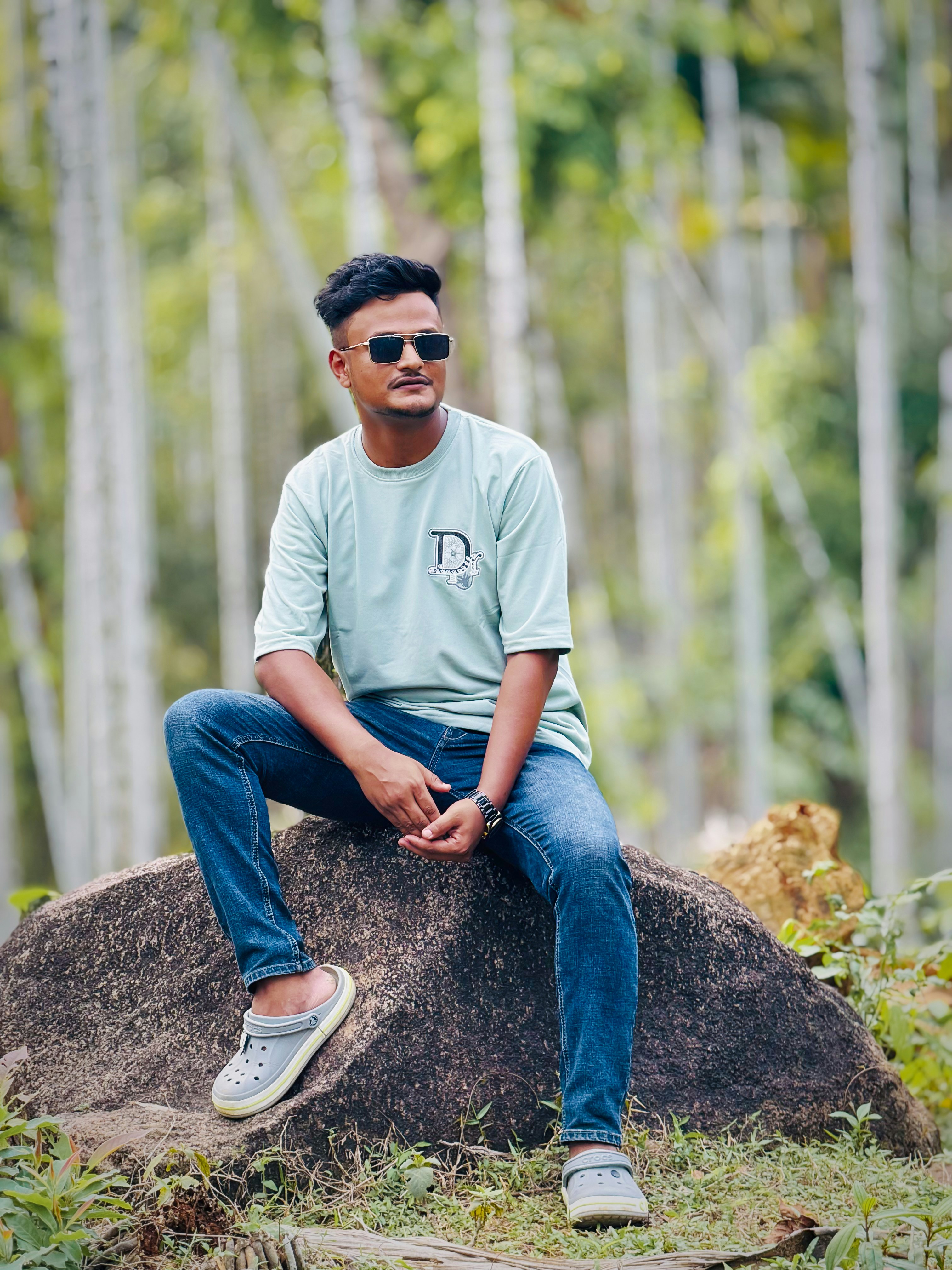 Young man in sunglasses sitting on a rock among trees.