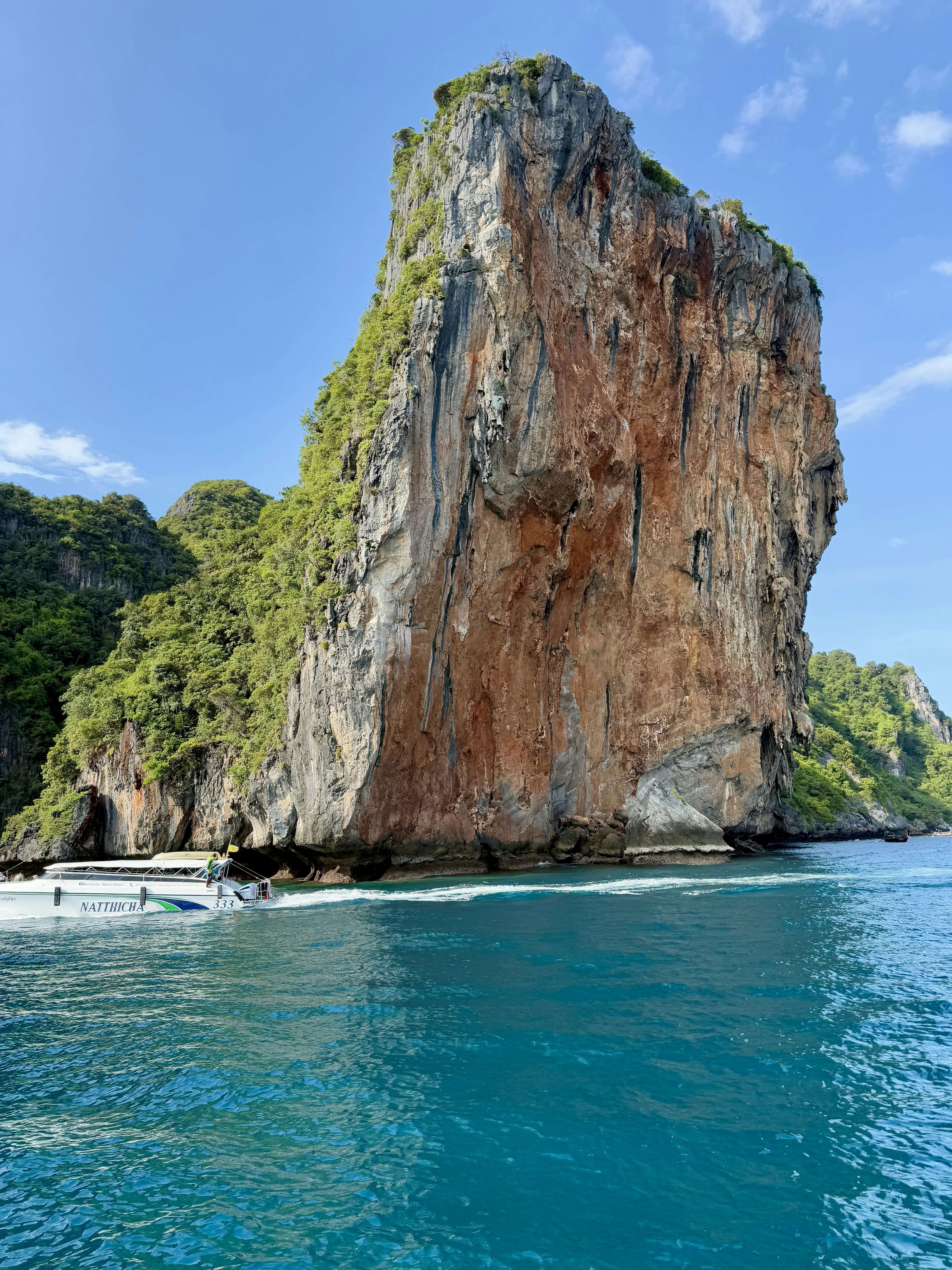 A towering limestone cliff rises dramatically above turquoise tropical waters beneath a clear blue sky.