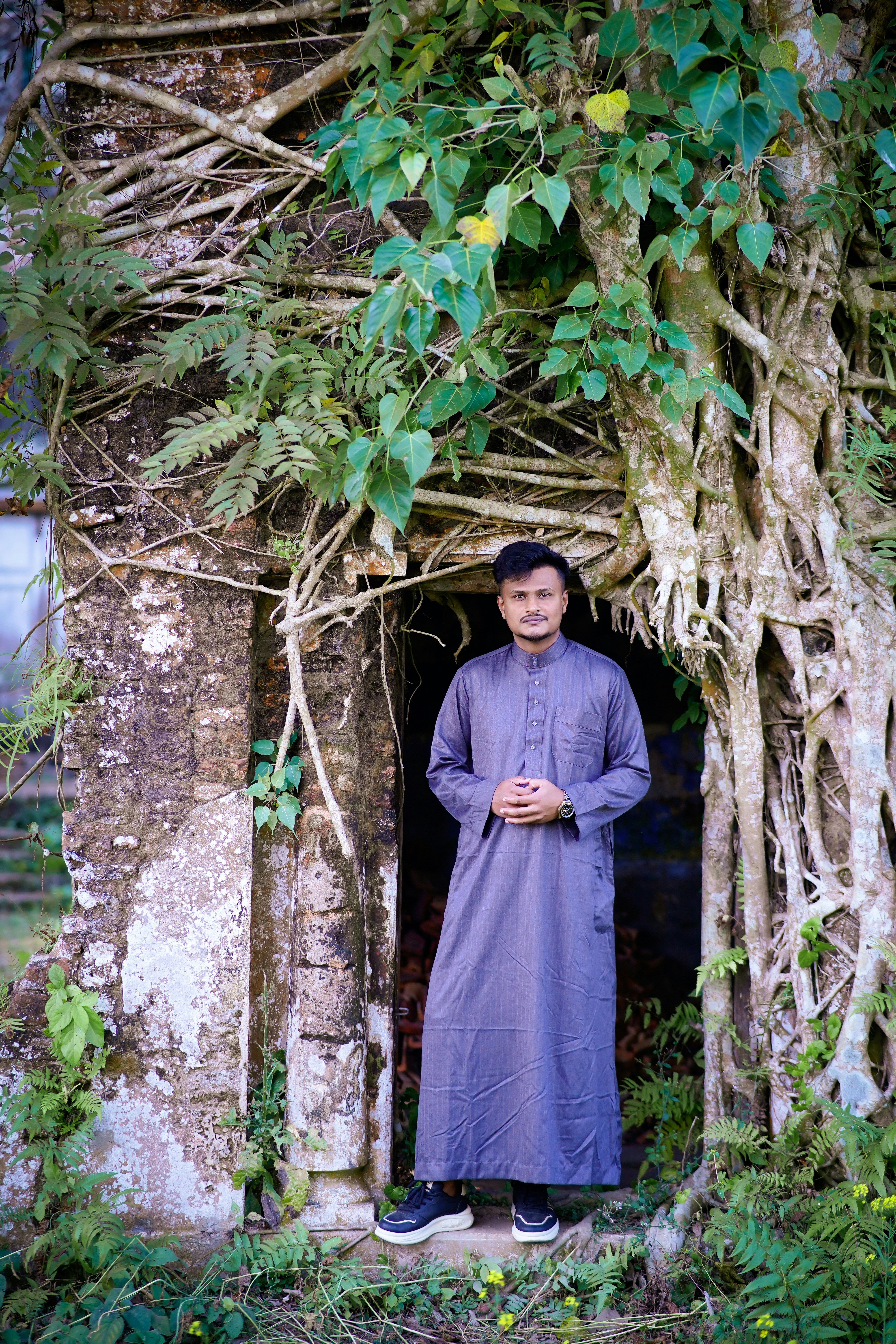 Man in a long gray tunic stands at the threshold of a vine-entwined doorway