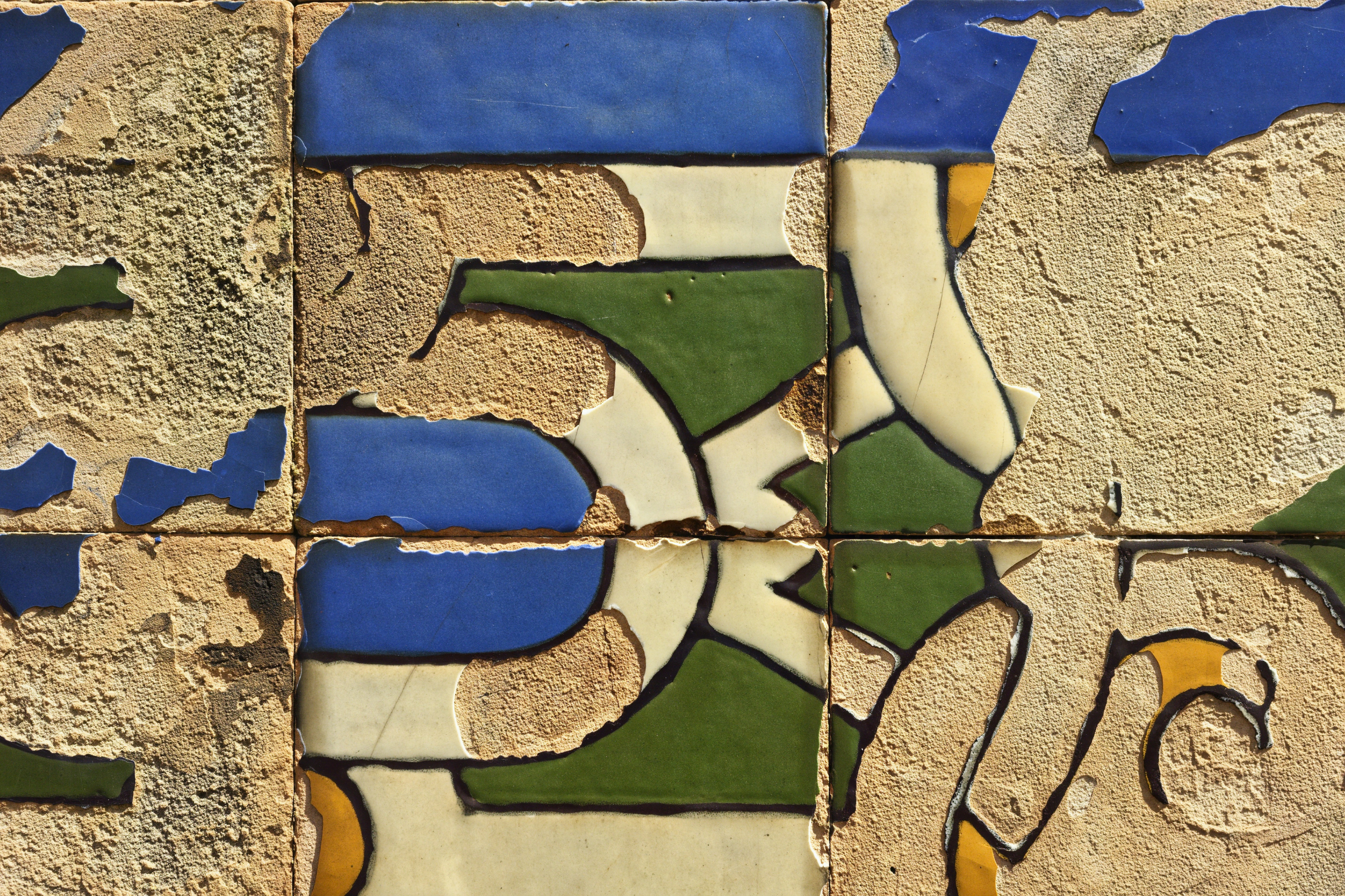 Decorative ceramic tiles on a wall with worn patterns and visible damage in a sunny outdoor setting - Colorful ceramic tiles show intricate patterns with some areas chipped and worn. Sunlight highlights the details of the design on a wall outdoors.