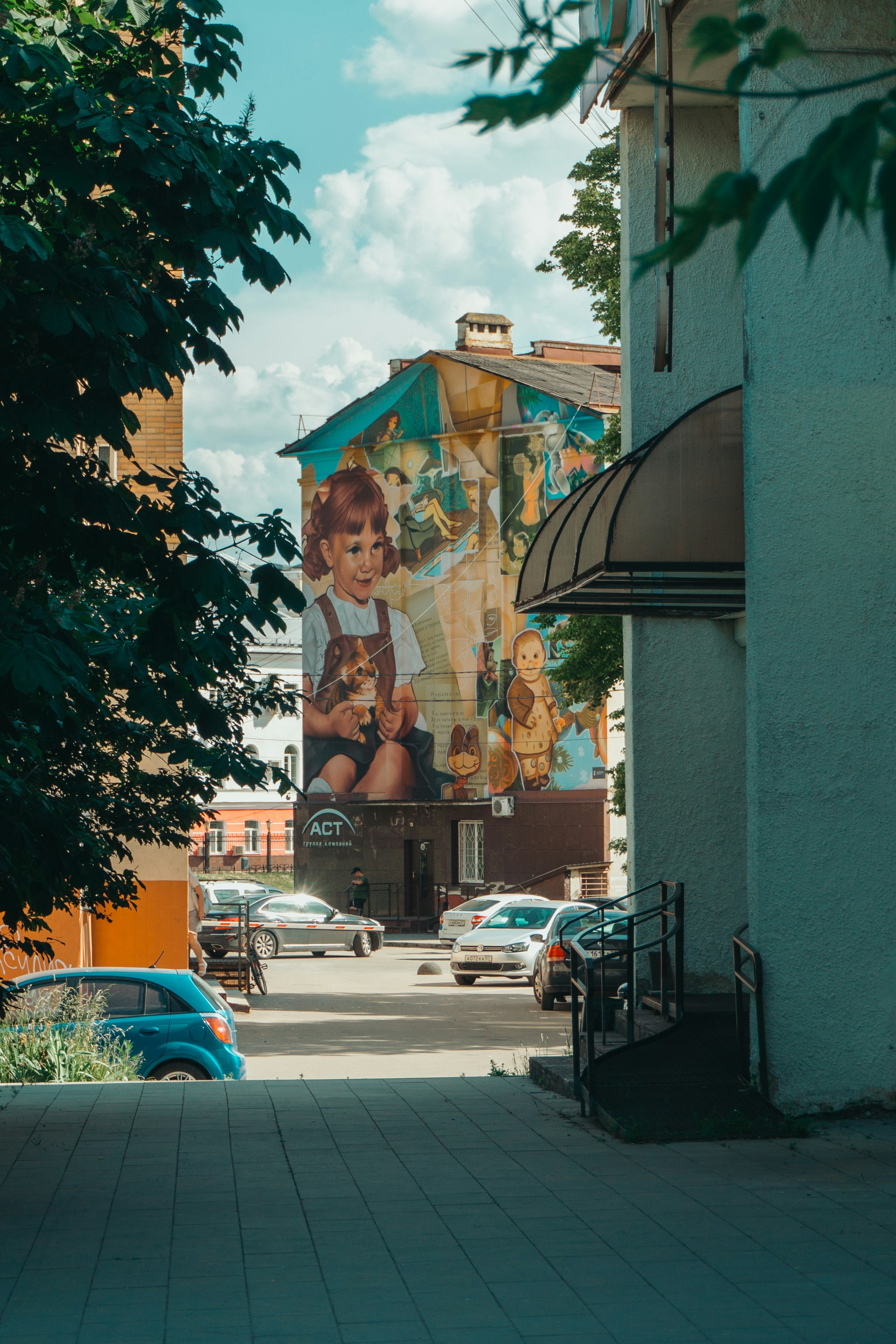 A large nostalgic mural of a girl with a cat covers the entire facade of a Soviet-era building, glimpsed through a narrow gap between city streets on a summer day.