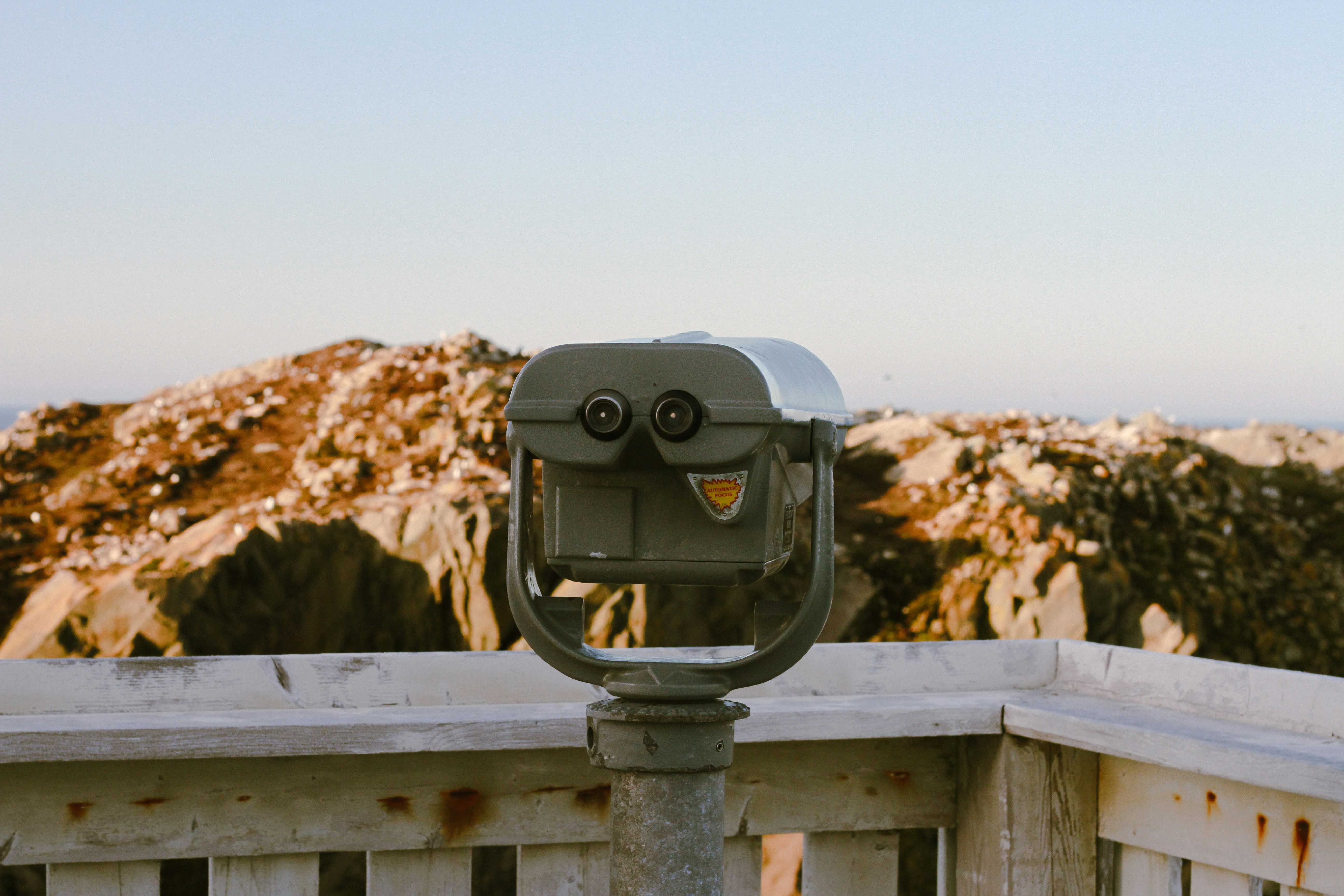 Close-range viewfinder perched on a weathered railing with rugged coastal cliffs in the background.