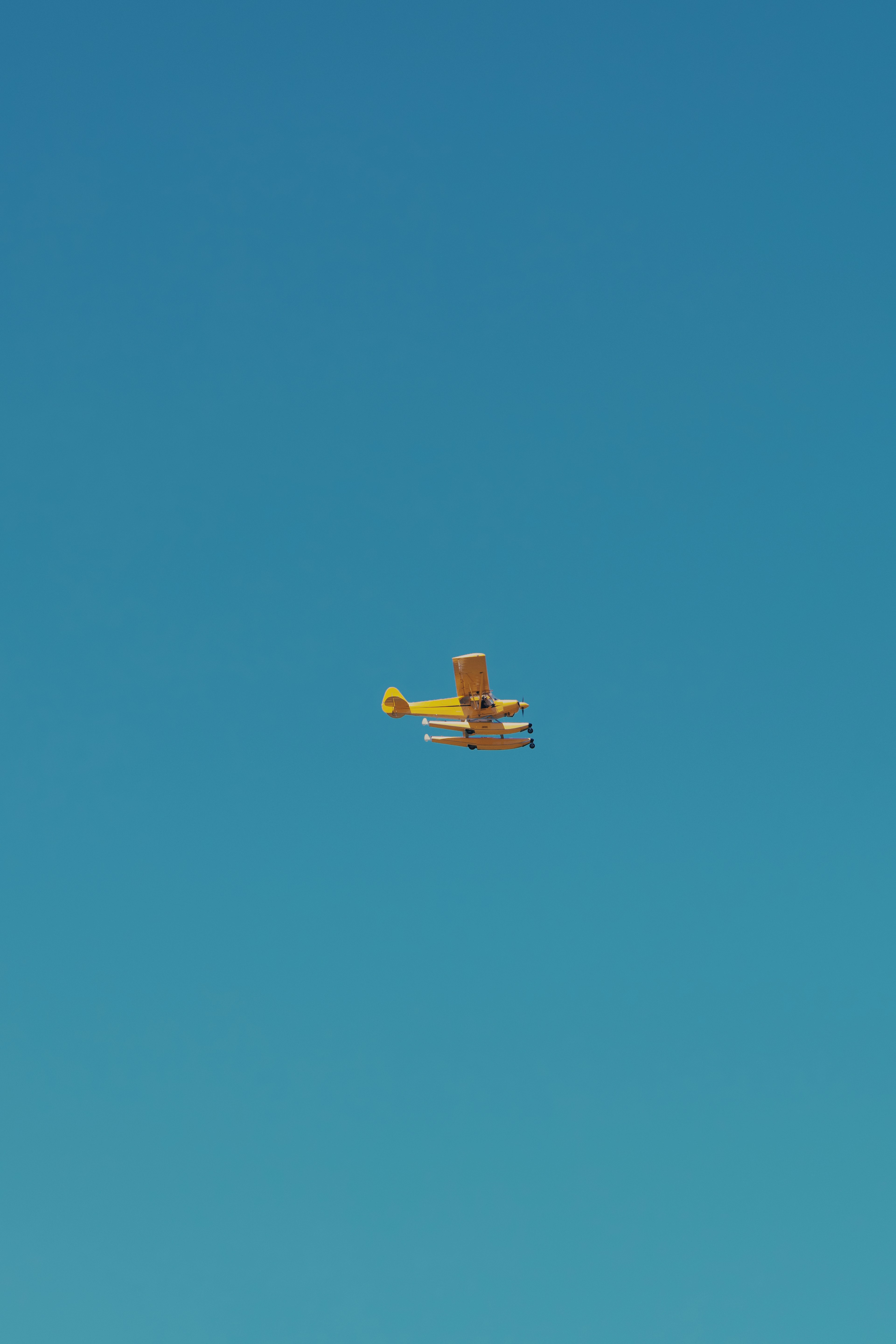A tiny yellow biplane seen in flight against a clear blue sky, captured with the aircraft centered in a wide open expanse.
