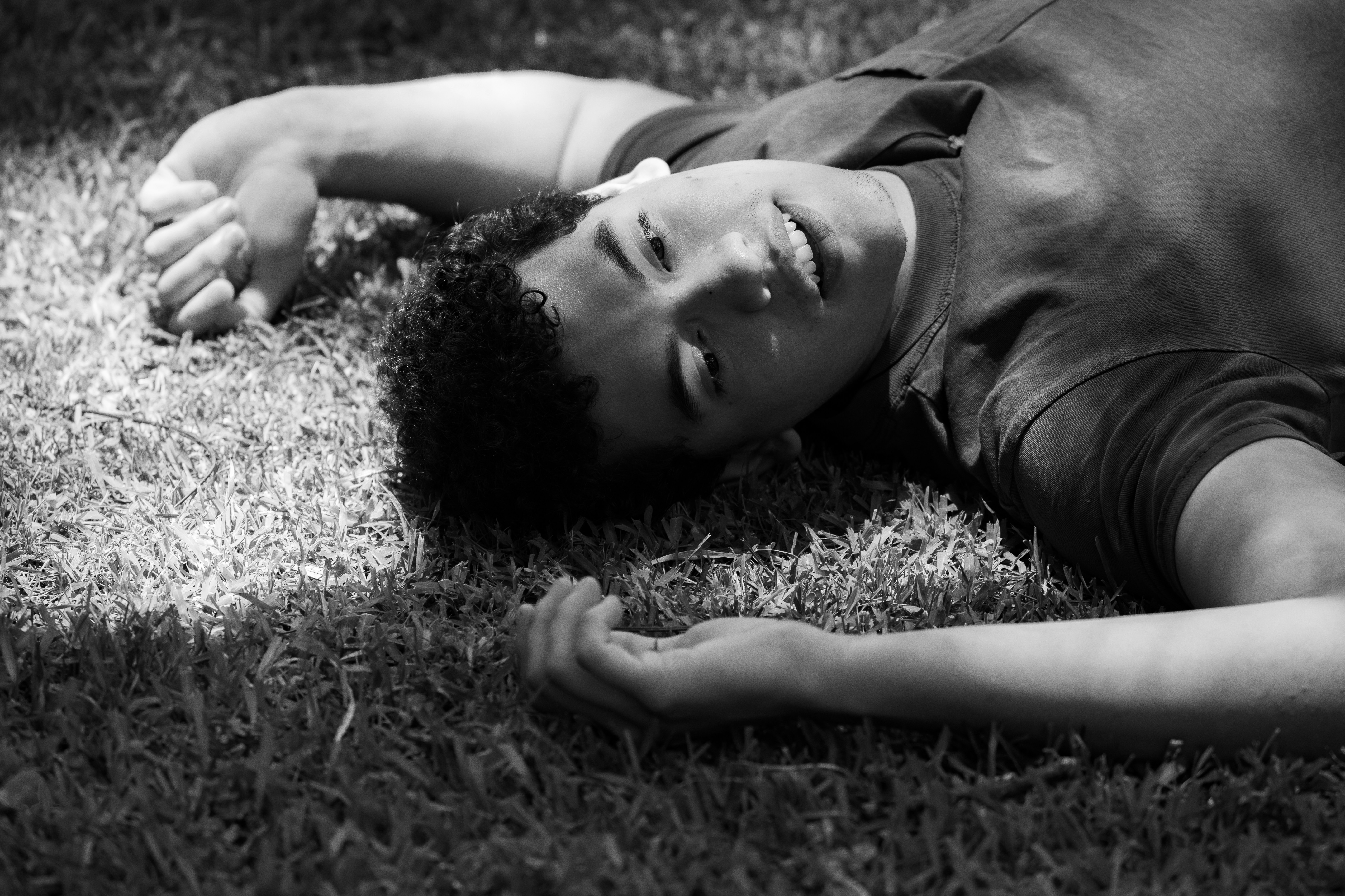 A relaxed young man lies on grass, face angled toward the camera, captured in black and white to emphasize mood and texture. A simple portrait focusing on expression and natural setting.