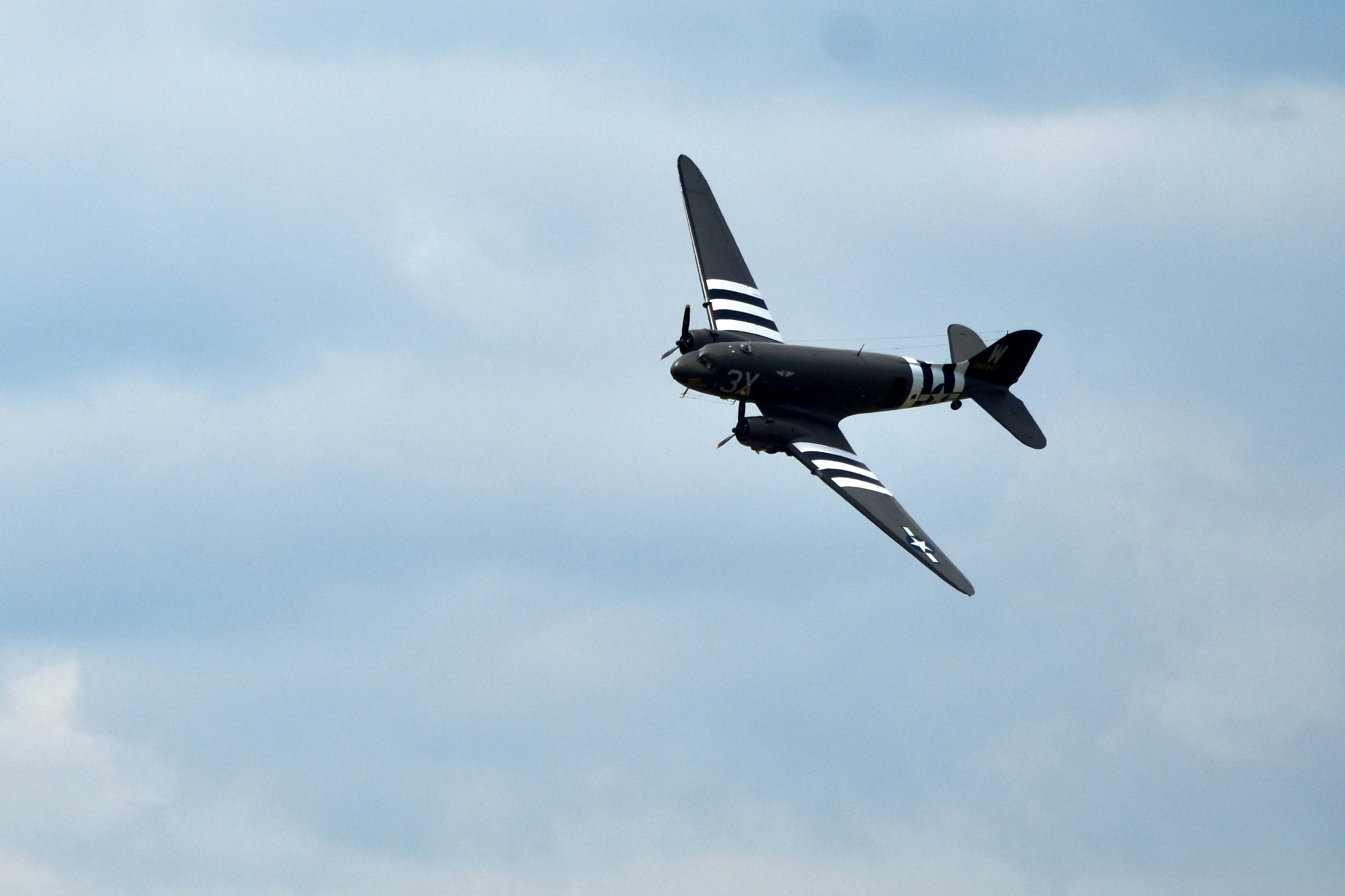 A black and white vintage propeller aircraft sails through a pale sky