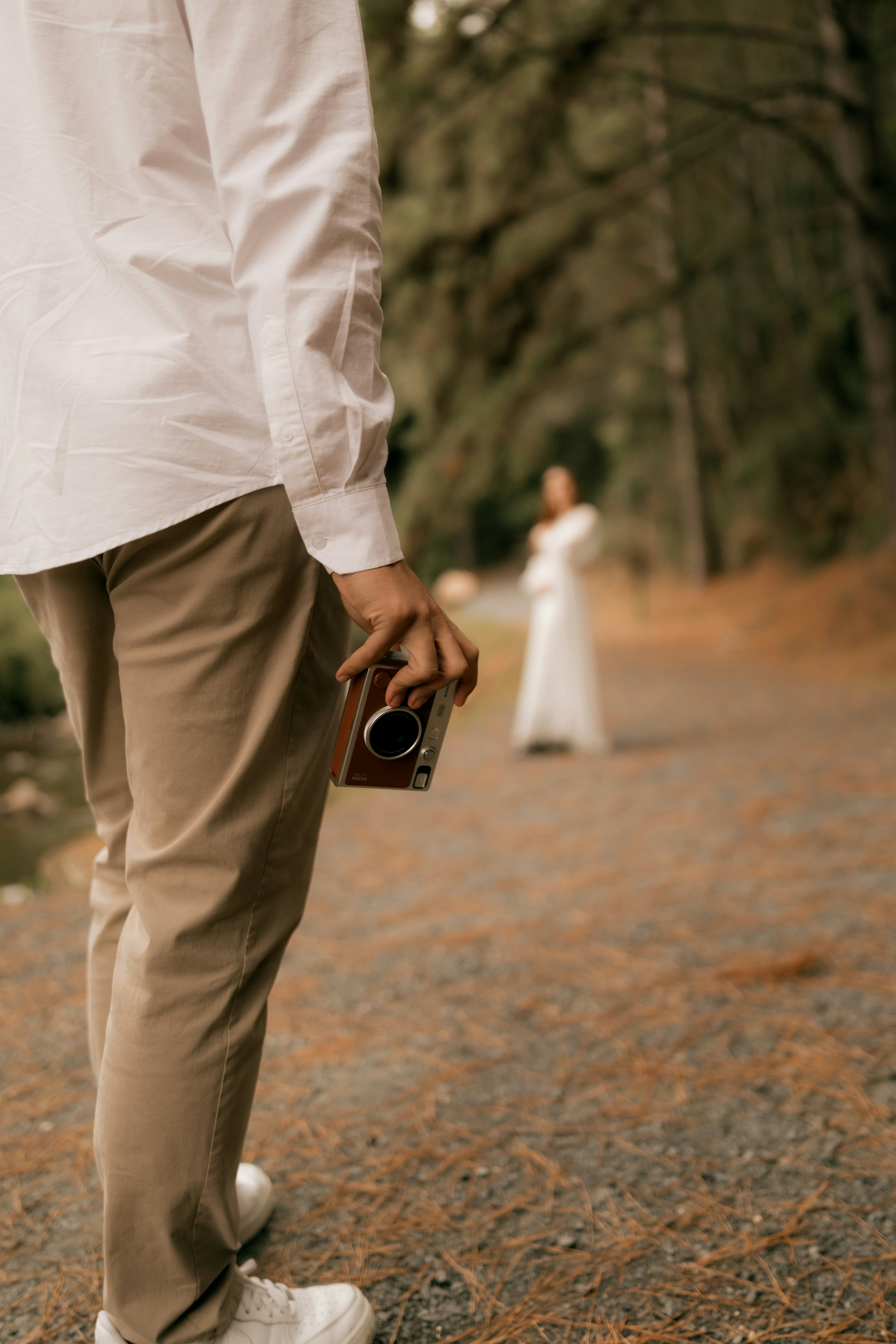 Man holding camera, woman in white dress walking on path.