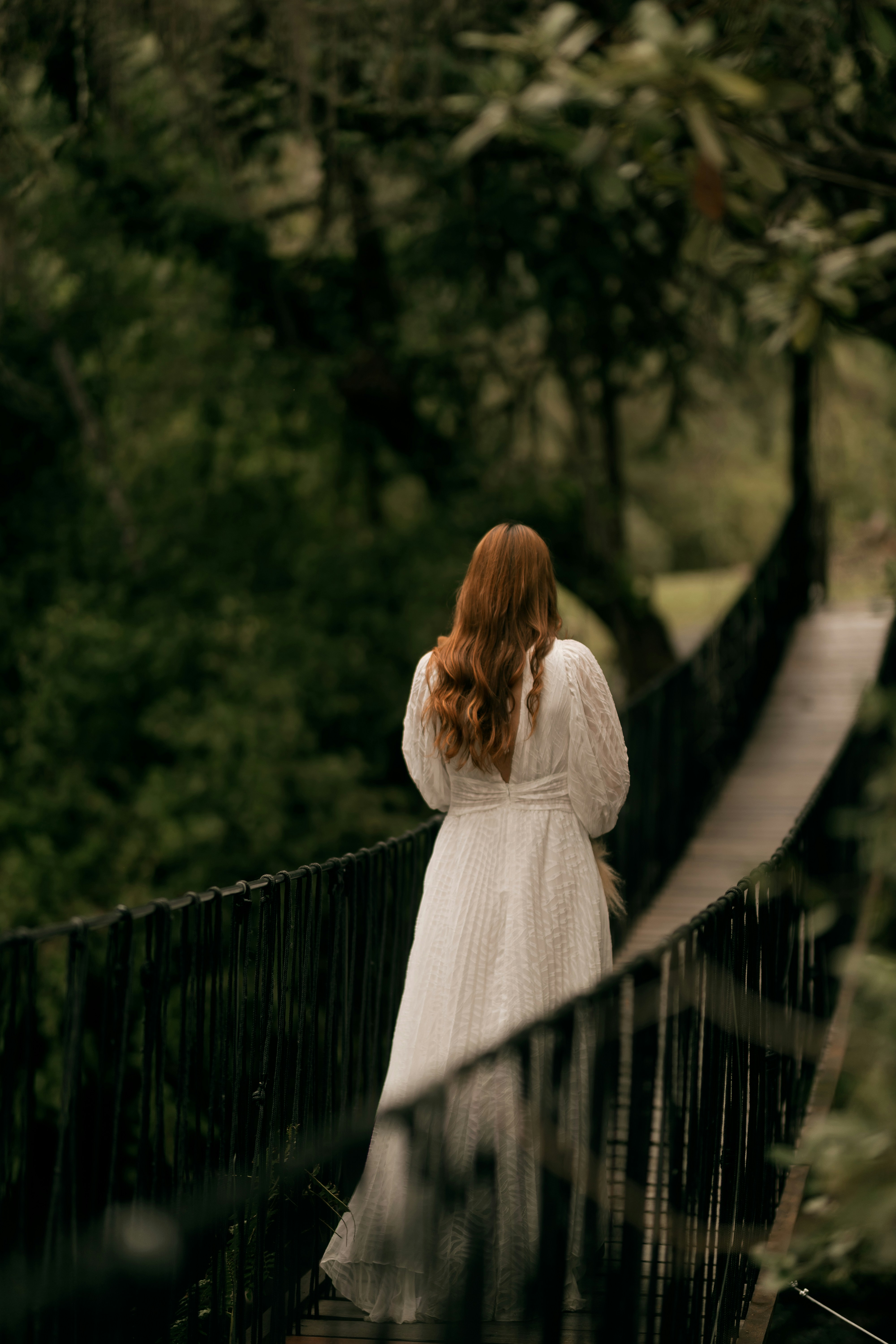 Red-haired woman in white dress walks on a forest bridge.