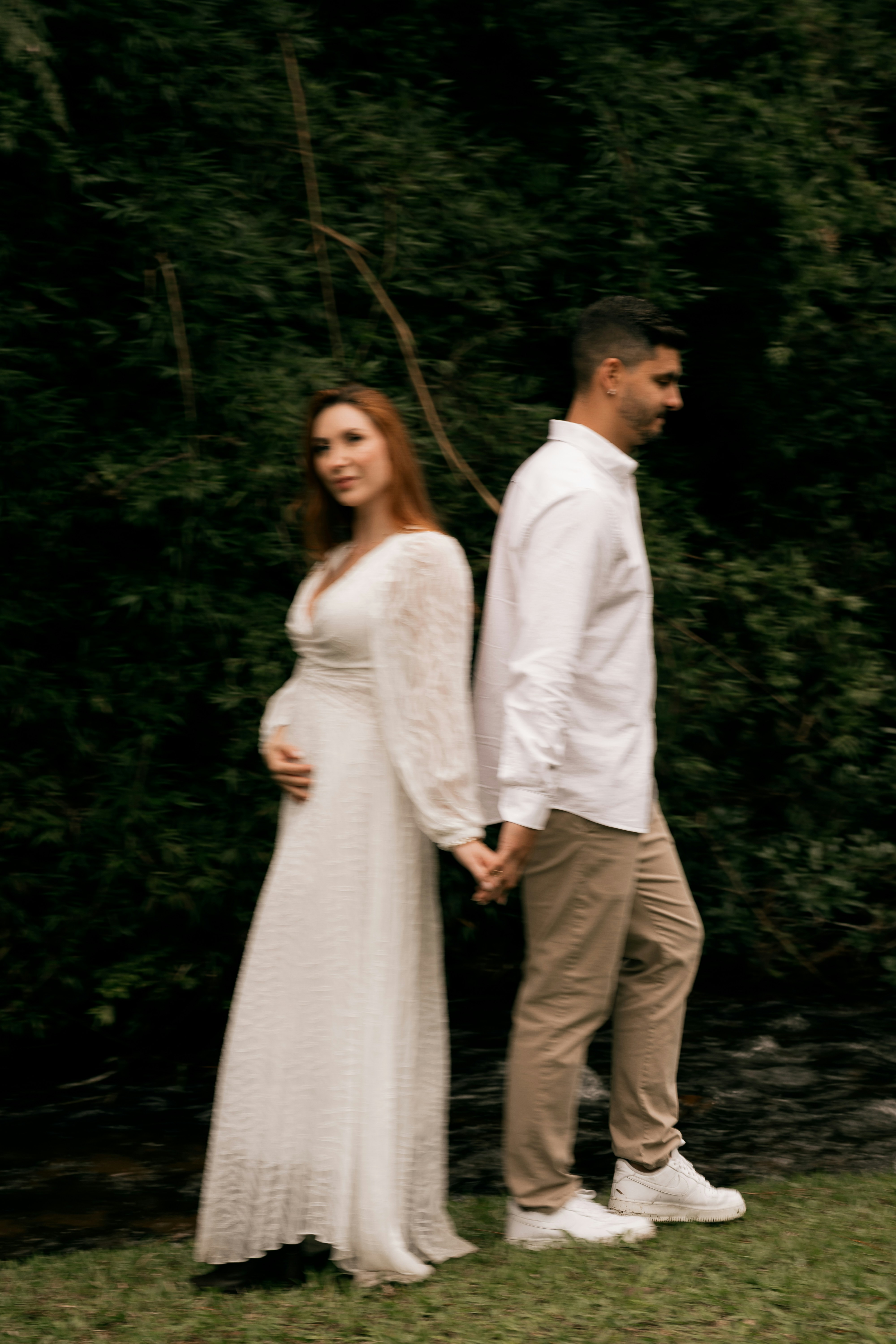 Expectant couple holding hands back-to-back in nature.