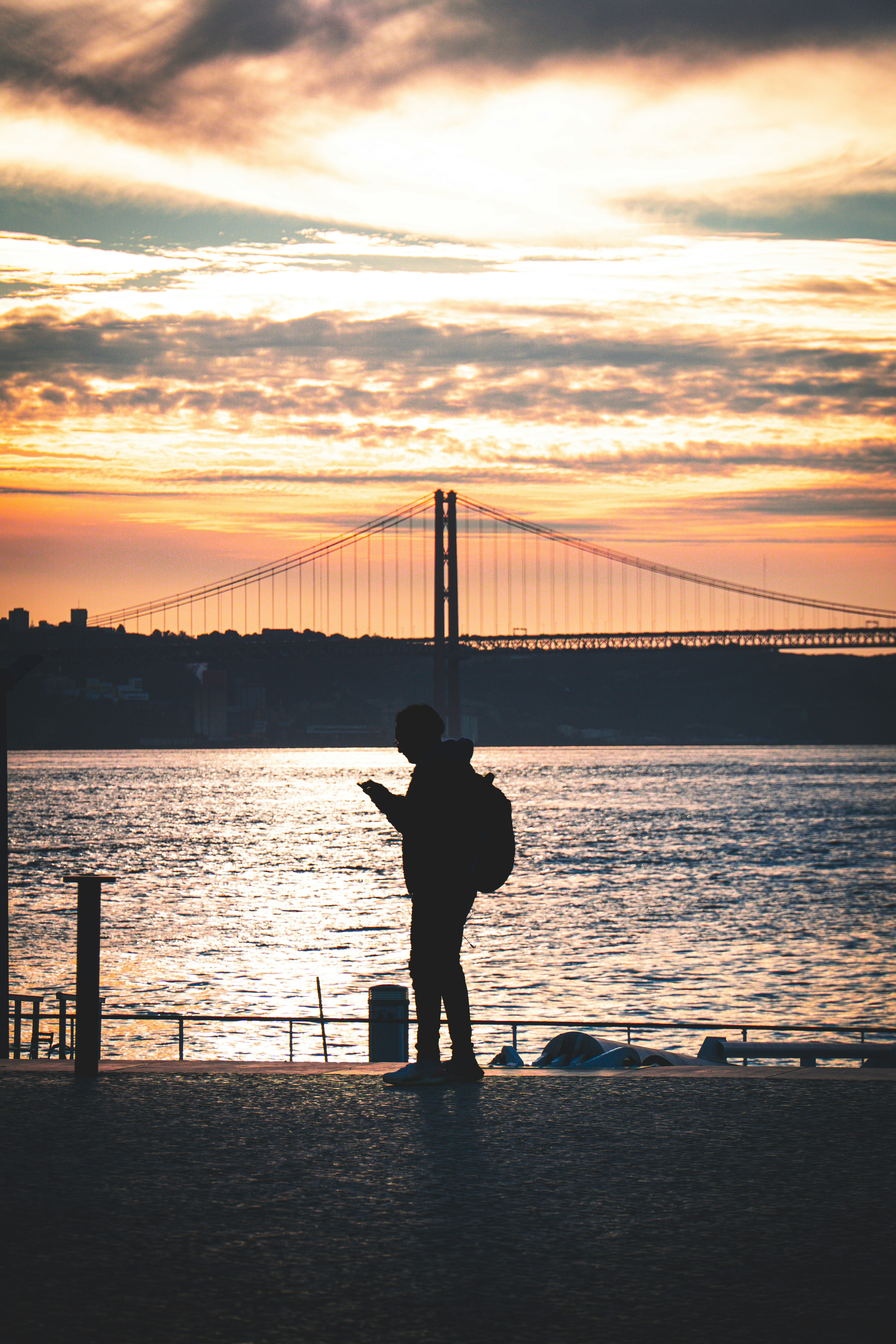 Sunset on the Tagus: A solitary figure stands along Lisbon’s waterfront as the setting sun illuminates the Tagus River and the bridge beyond.