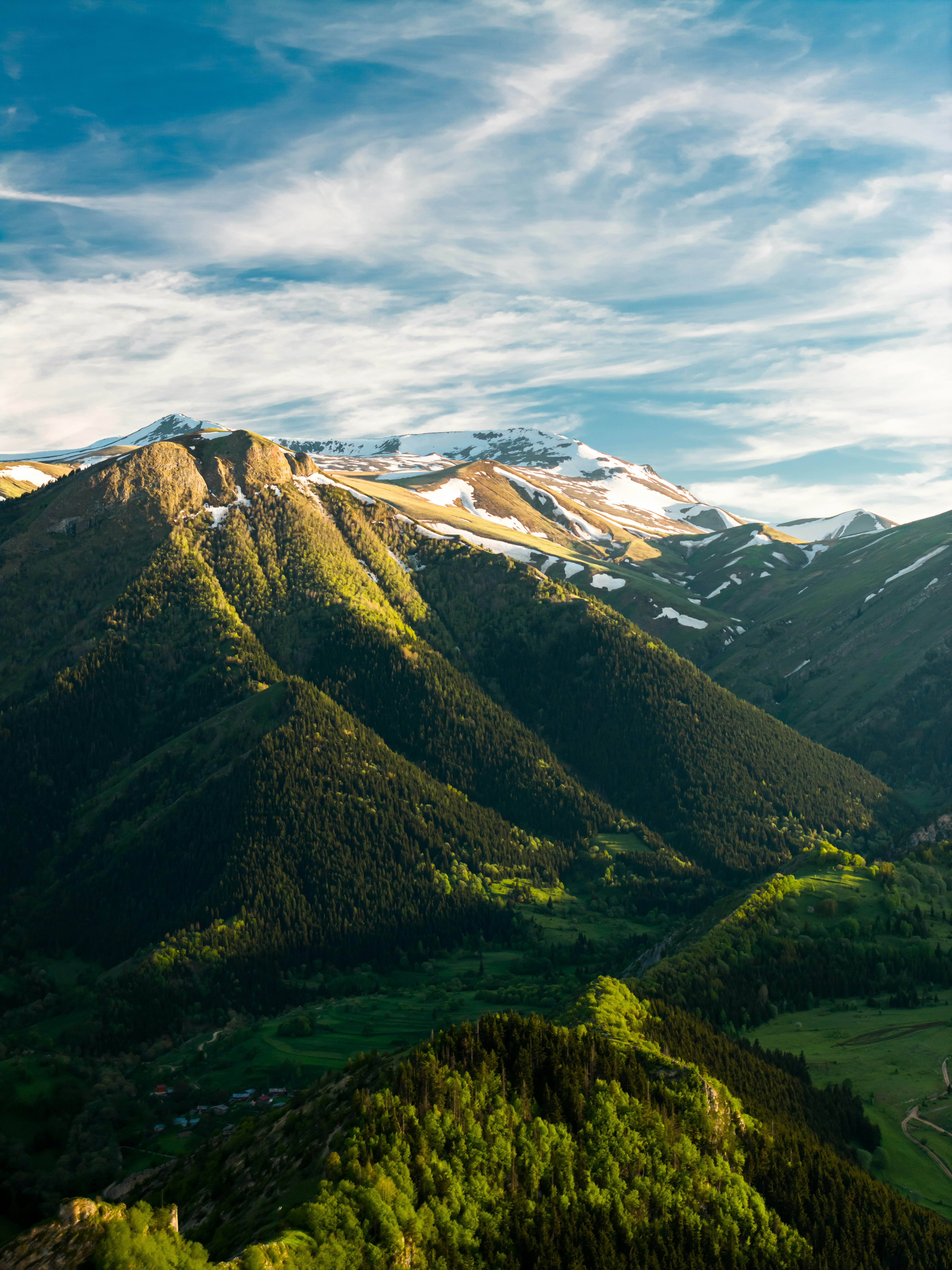 Snow-capped mountains and green forests under a cloudy blue sky.