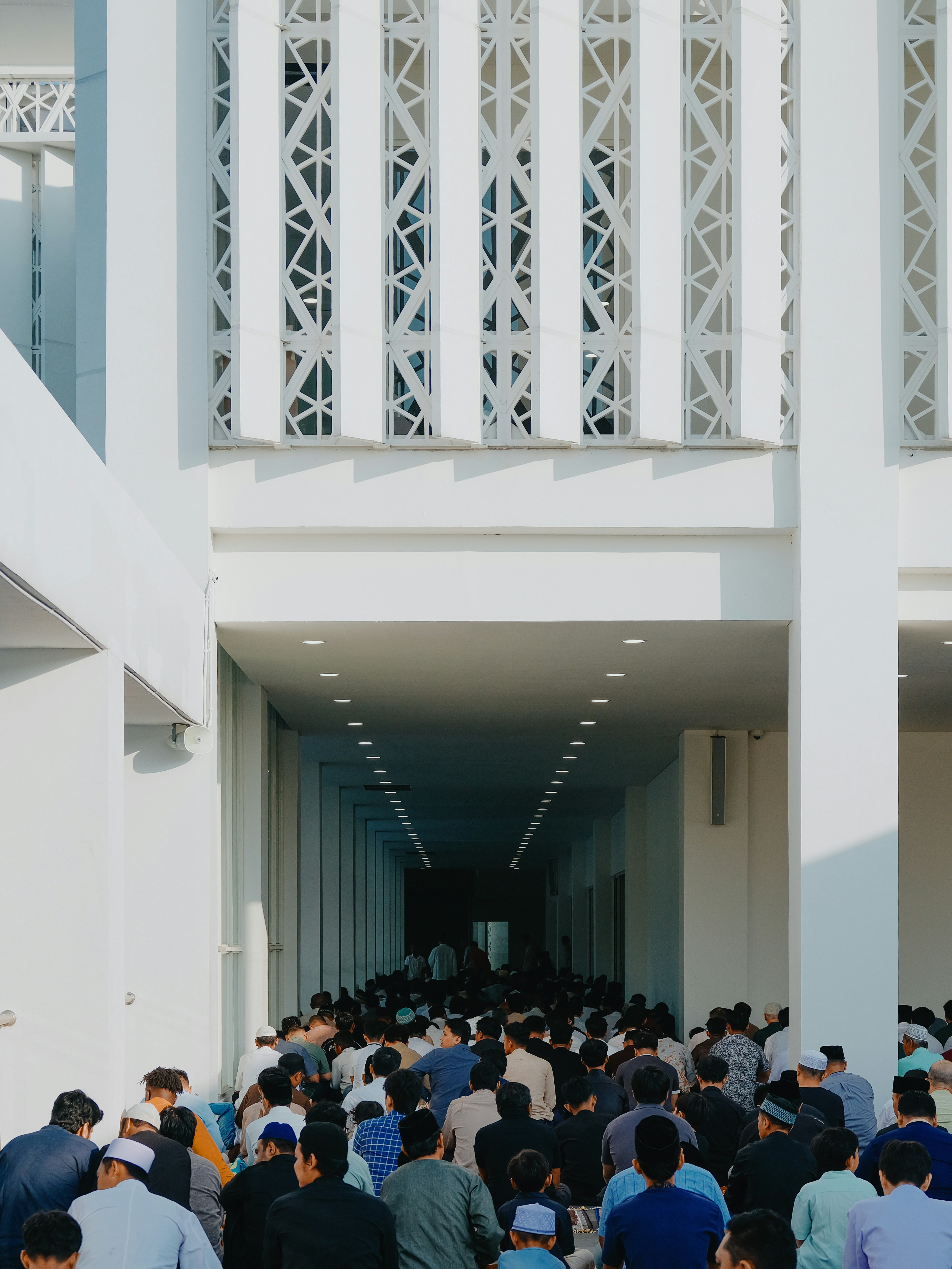 Worshippers gather and sit for Eid al-Adha prayer at the end of a bright corridor at Masjid Raya Baitul Mukhtar, BSD City, framed by the mosque's white geometric lattice screens.