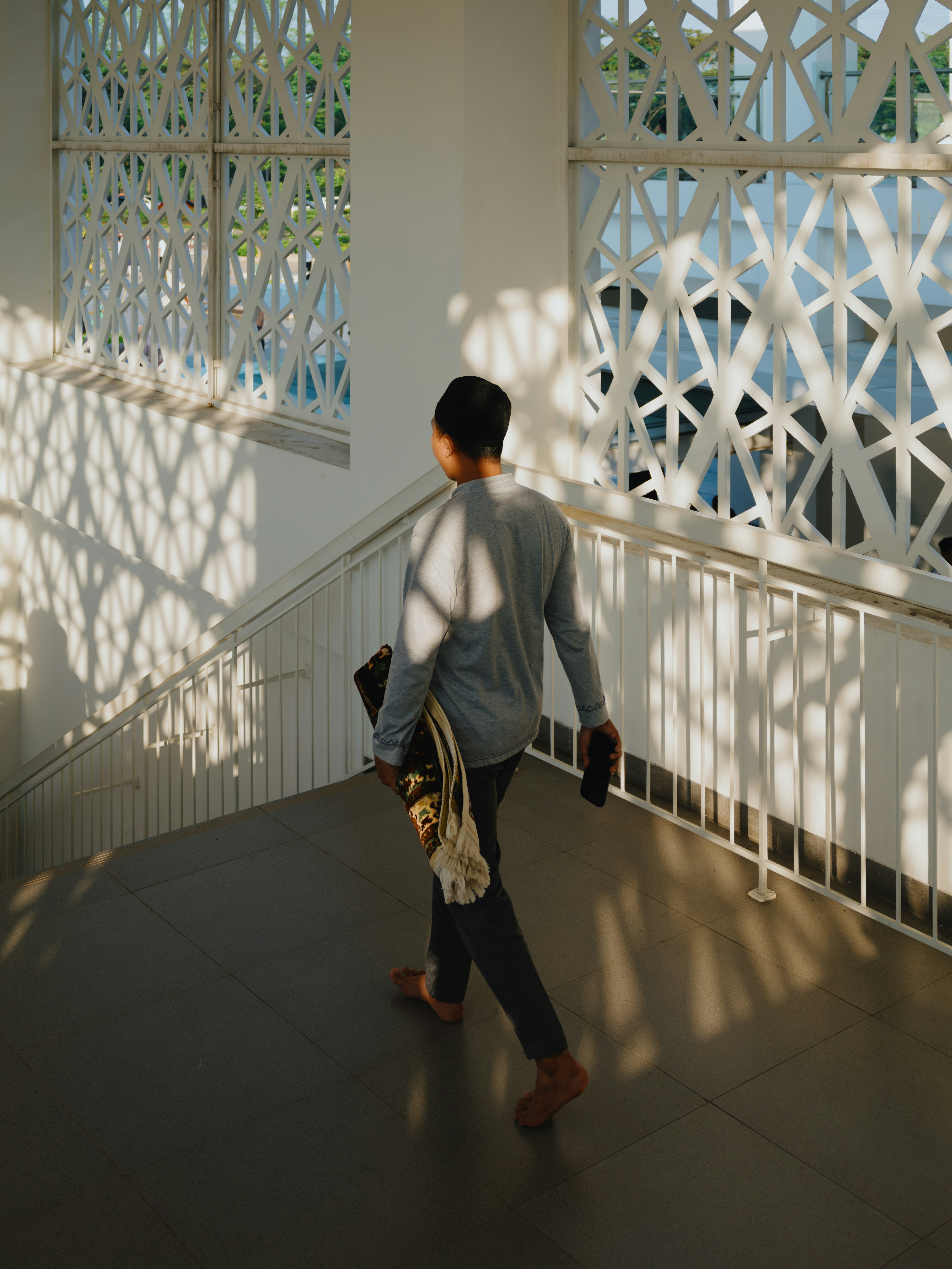 A man in a grey shirt holding a prayer mat and his phone walks down the staircase at Masjid Raya Baitul Mukhtar, BSD City, lit by sunlight passing through the geometric lattice during Eid al-Adha.