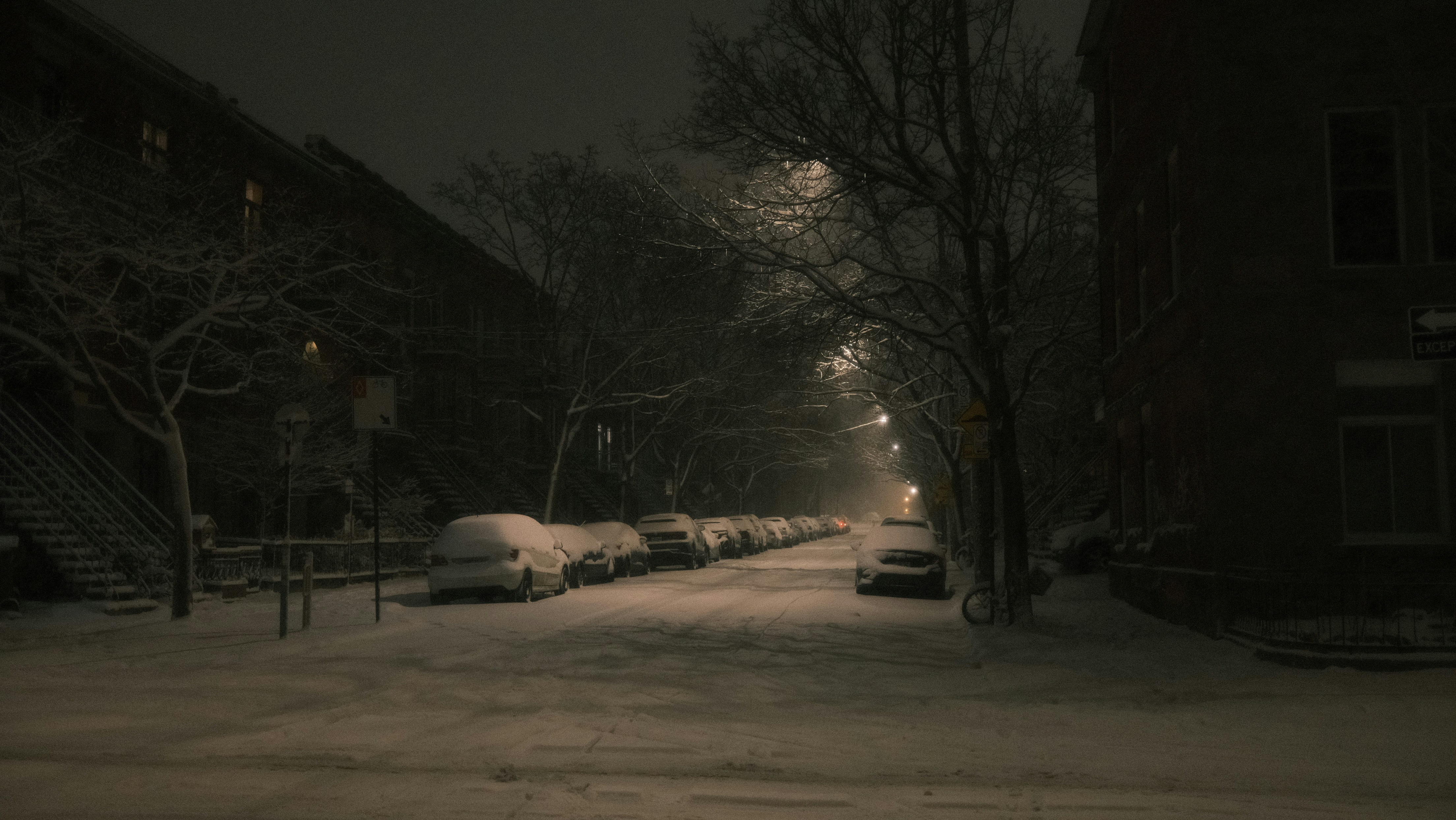 Snow-covered street with parked cars under dim streetlights at night.
