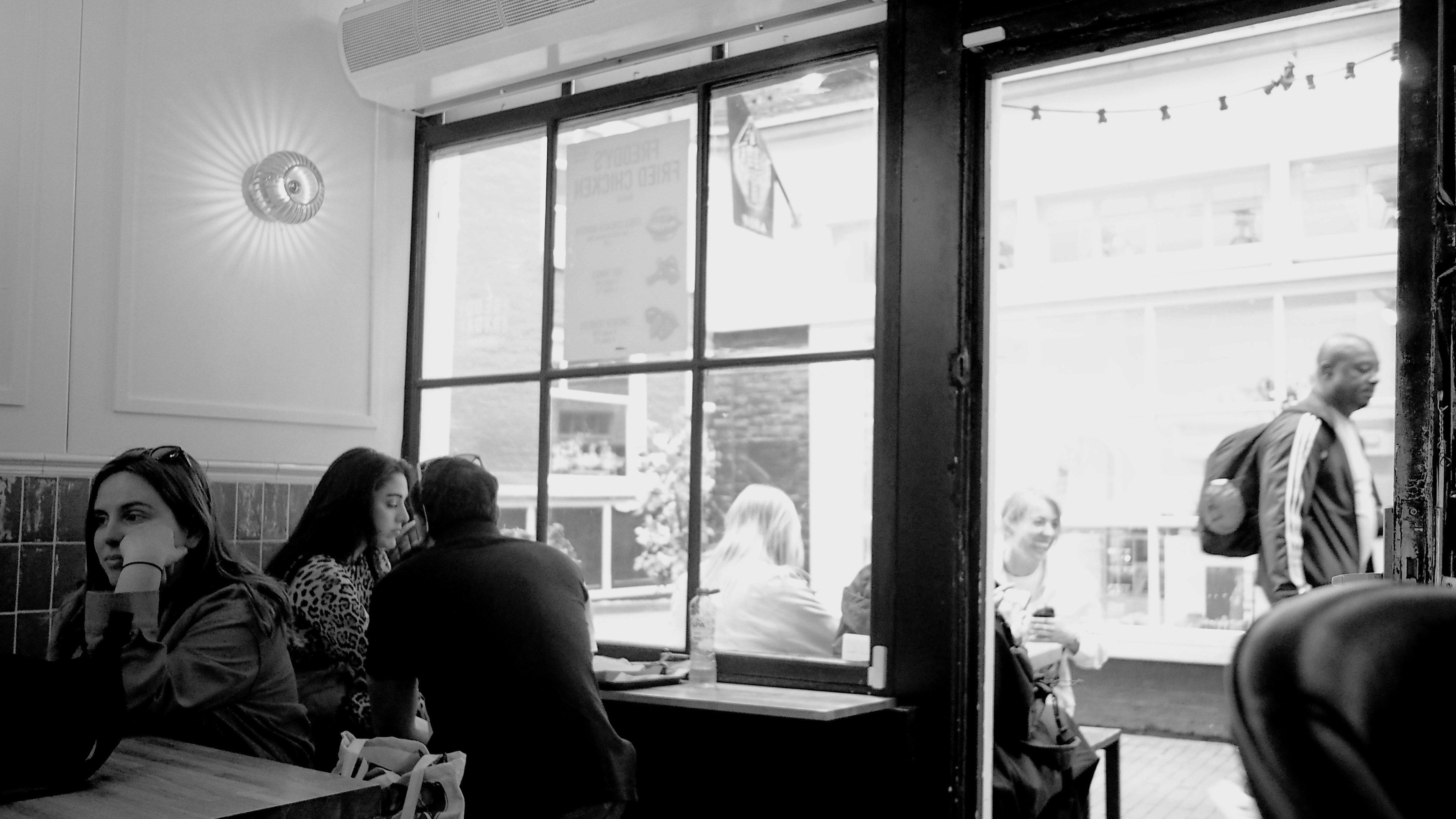 A moody, high-contrast monochrome photograph capturing a quiet, candid moment inside a busy snackbar in Amsterdam, Netherlands. A group of patrons sits at a large window table, their forms in partial silhouette and shadow, looking out onto a narrow street. Beyond the glass, other people are visible walking, providing a sense of street-level movement and life. In the foreground, an ornate sunburst wall sconce glows against the clean interior wall.