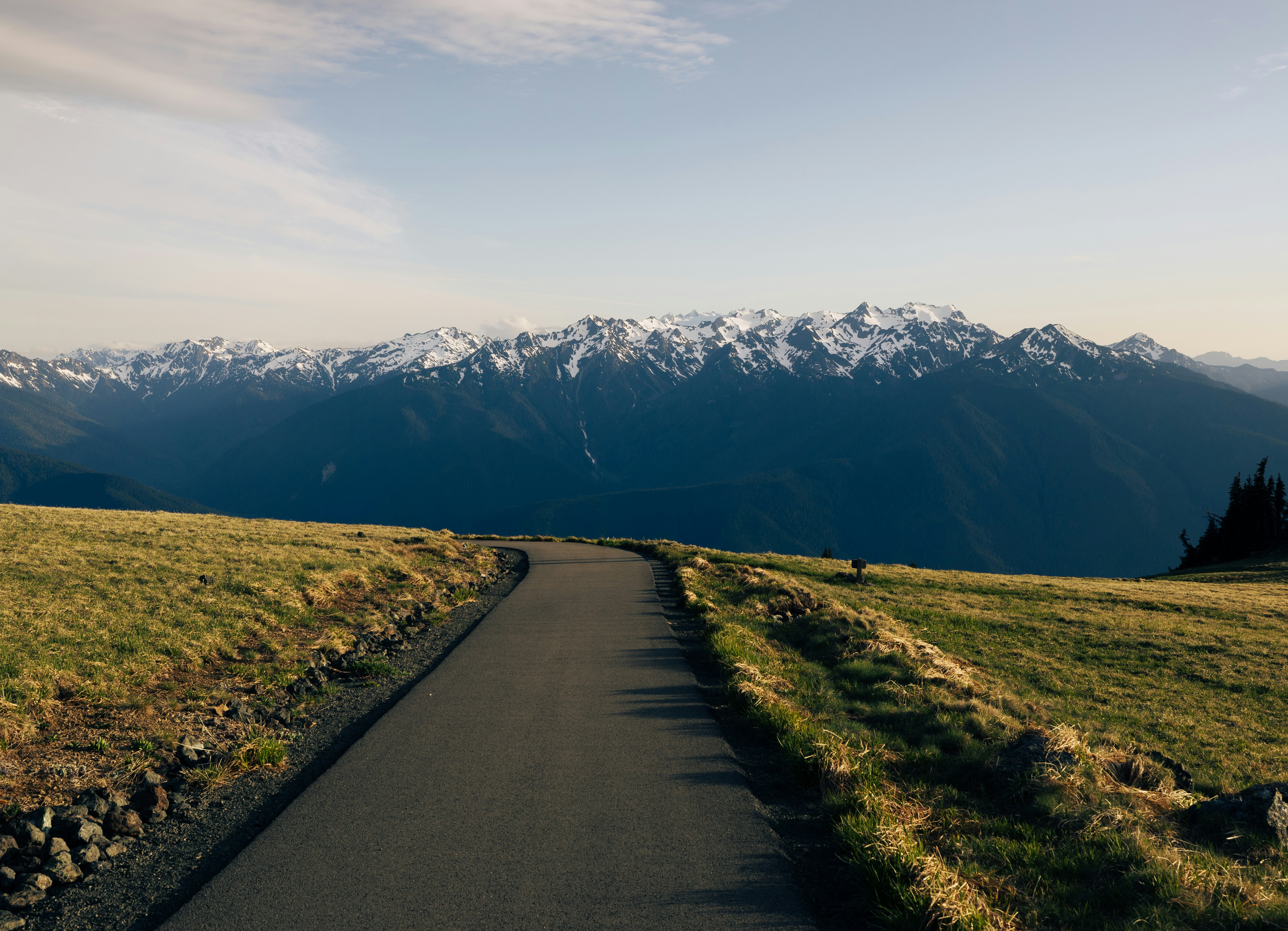 Winding mountain road leads toward snow-capped peaks under a clear sky, framed by grassy shoulders. This landscape shot highlights depth and sense of journey.