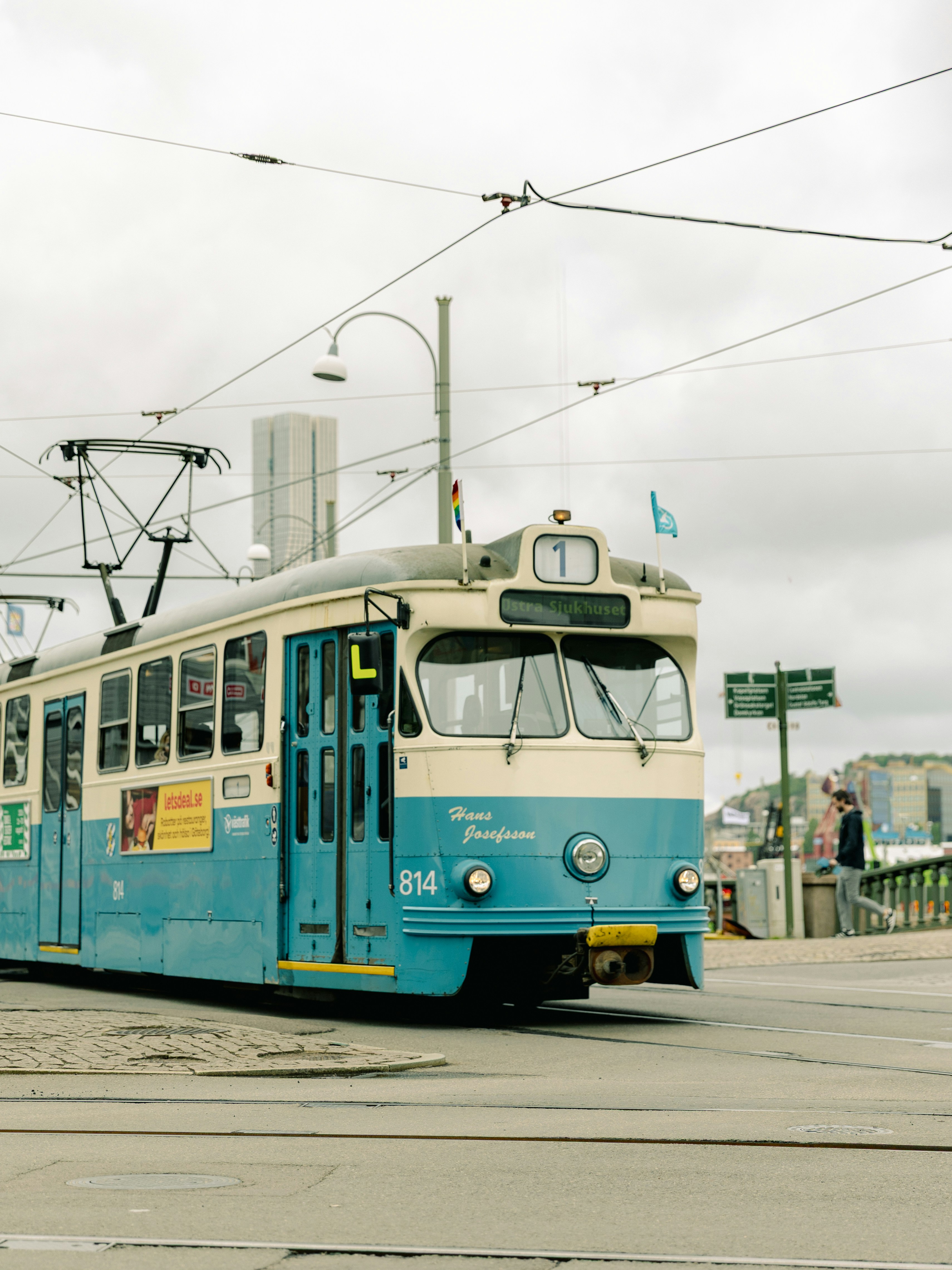 Blue tram with upper deck, crossing a street under overhead wires; urban backdrop with distant buildings and a pedestrian on the platform.