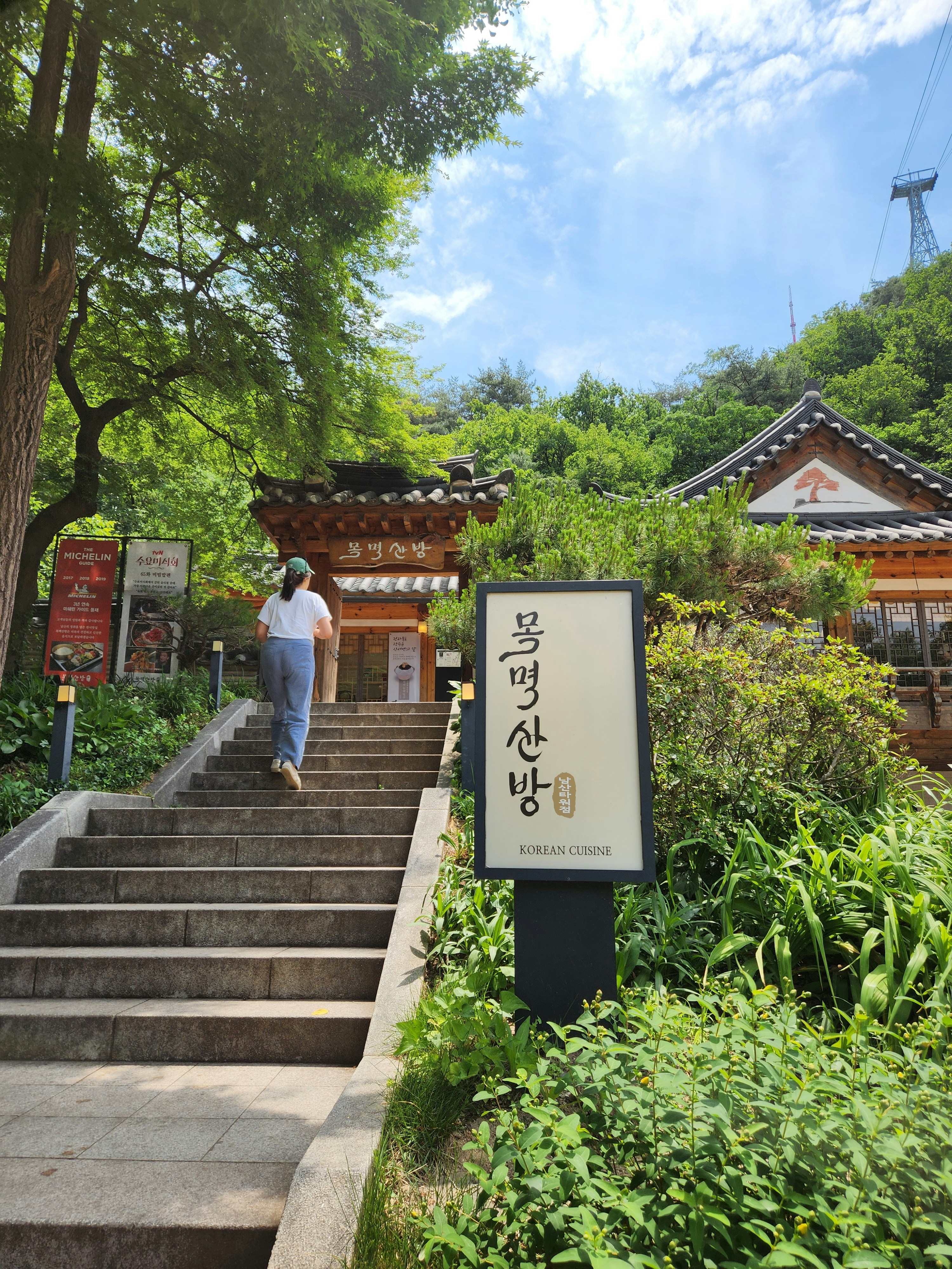 Stone steps lead to a traditional wooden gate amid lush greenery; a person ascends toward the entrance, while a vertical sign and surrounding garden frame the scene. A Korean cuisine sign suggests a restaurant interior beyond.