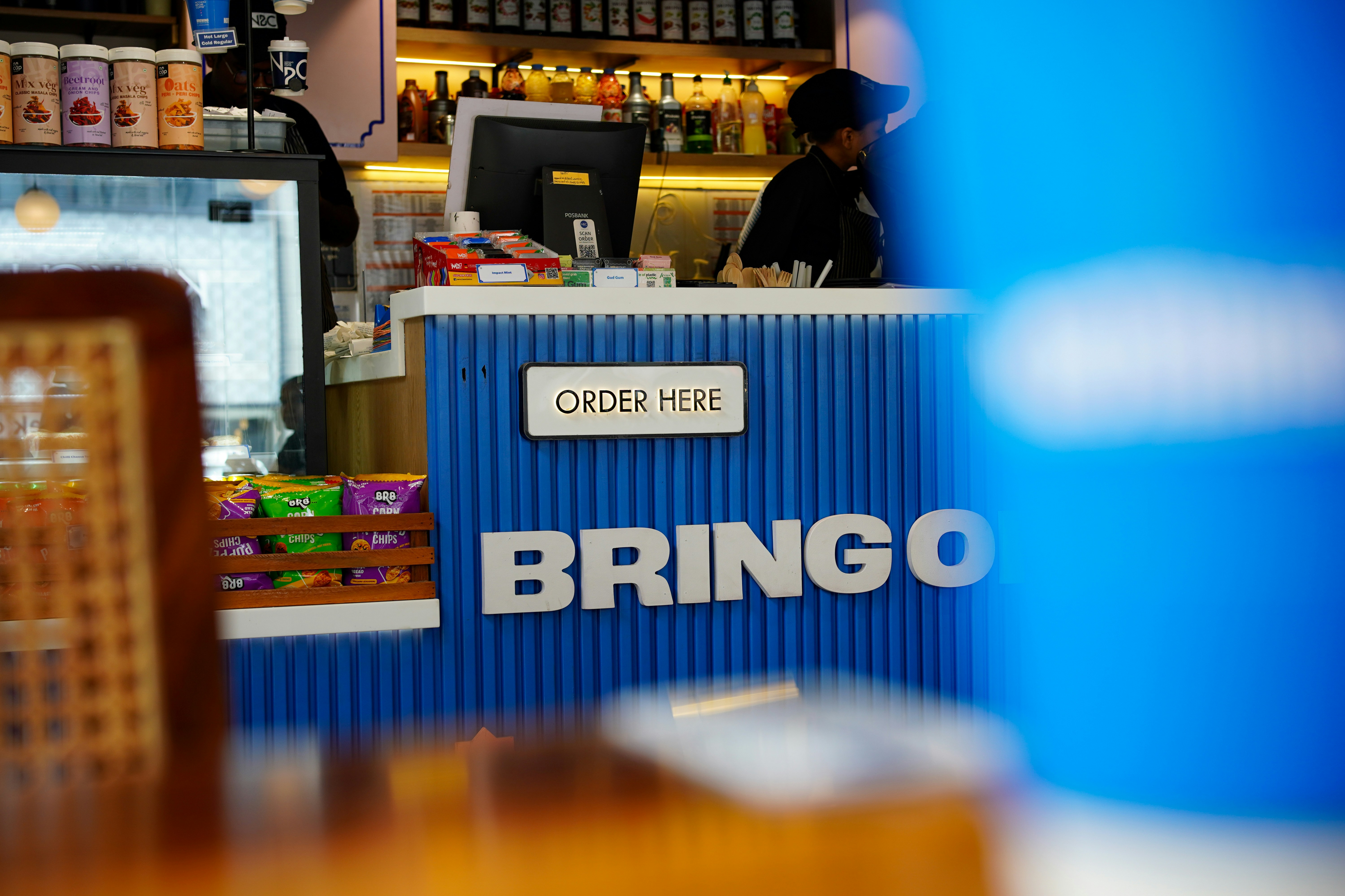 A candid shot of a modern cafe counter with a prominent "ORDER HERE" sign, capturing the warm, inviting atmosphere of a bustling coffee shop. The composition uses a shallow depth of field to draw focus to the service point while maintaining the authentic, busy character of the workspace.