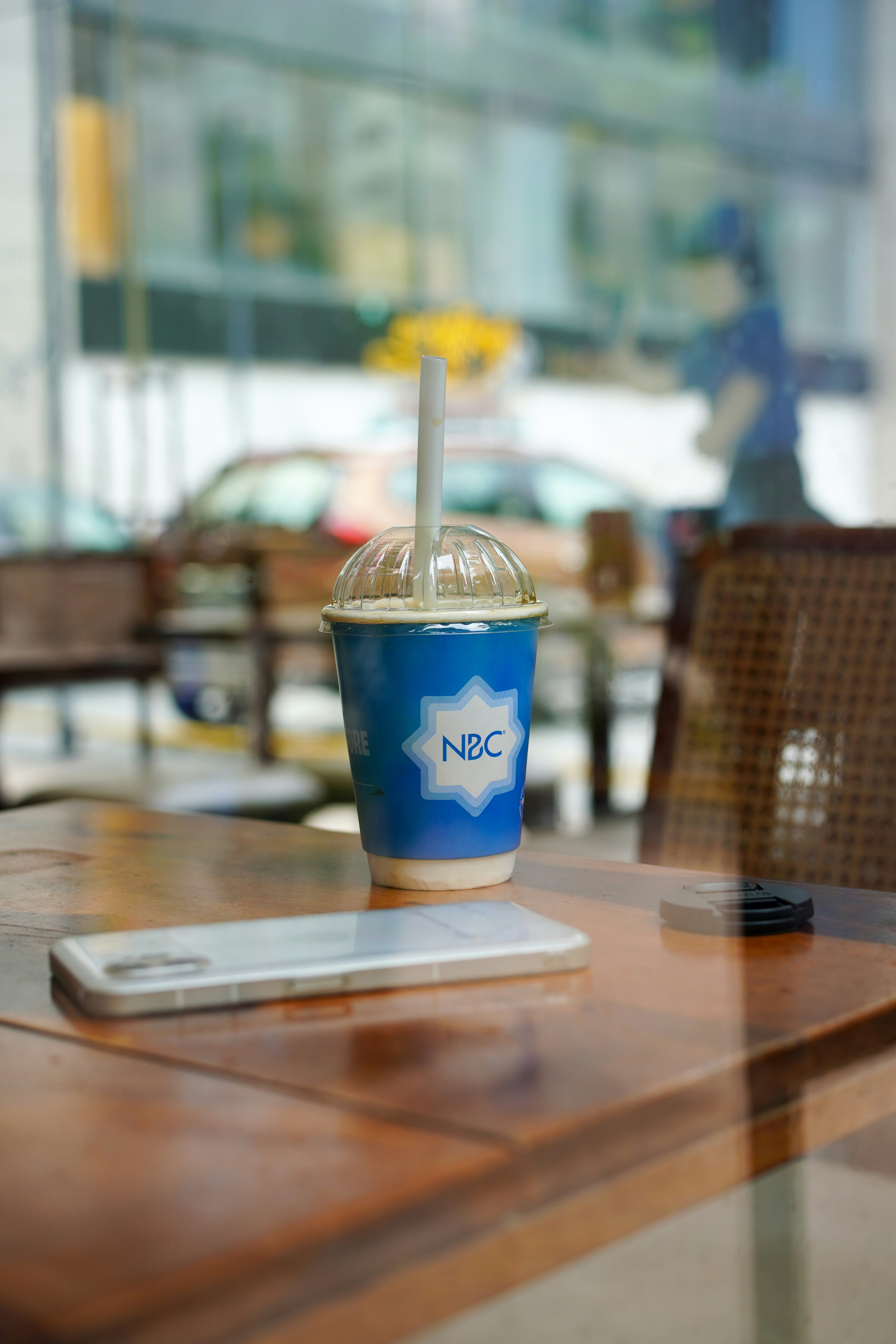 A relaxed scene featuring a branded blue coffee cup and a smartphone resting on a wooden cafe table. The shallow depth of field frames the subject against a softly blurred city window view, perfectly capturing a quiet, modern coffee shop moment.
