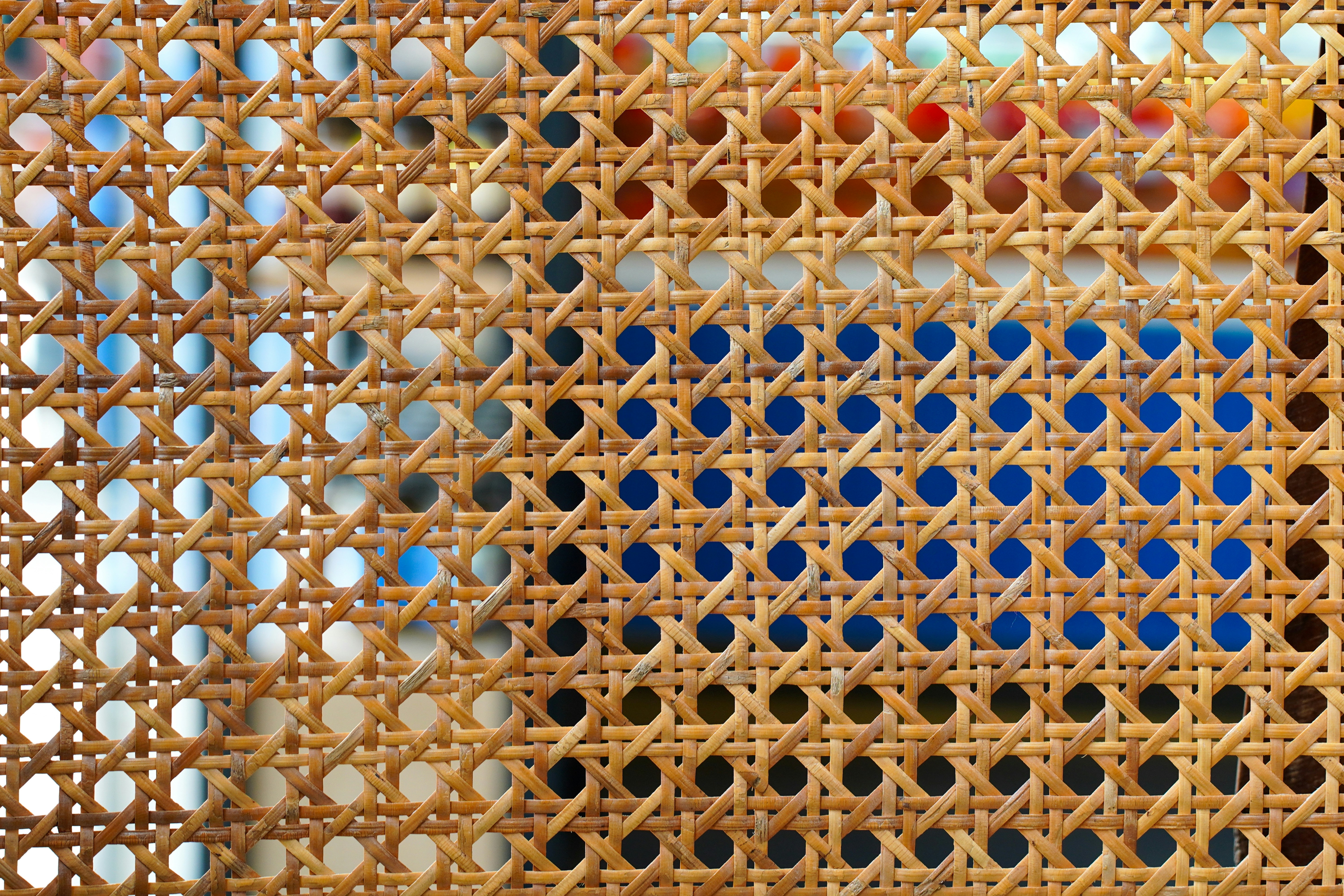 A close-up photograph featuring the classic, repetitive weave of a cane furniture surface. The natural warmth of the material contrasts subtly with glimpses of a colorful, blurred background, highlighting the intersection of traditional craftsmanship and modern decor.