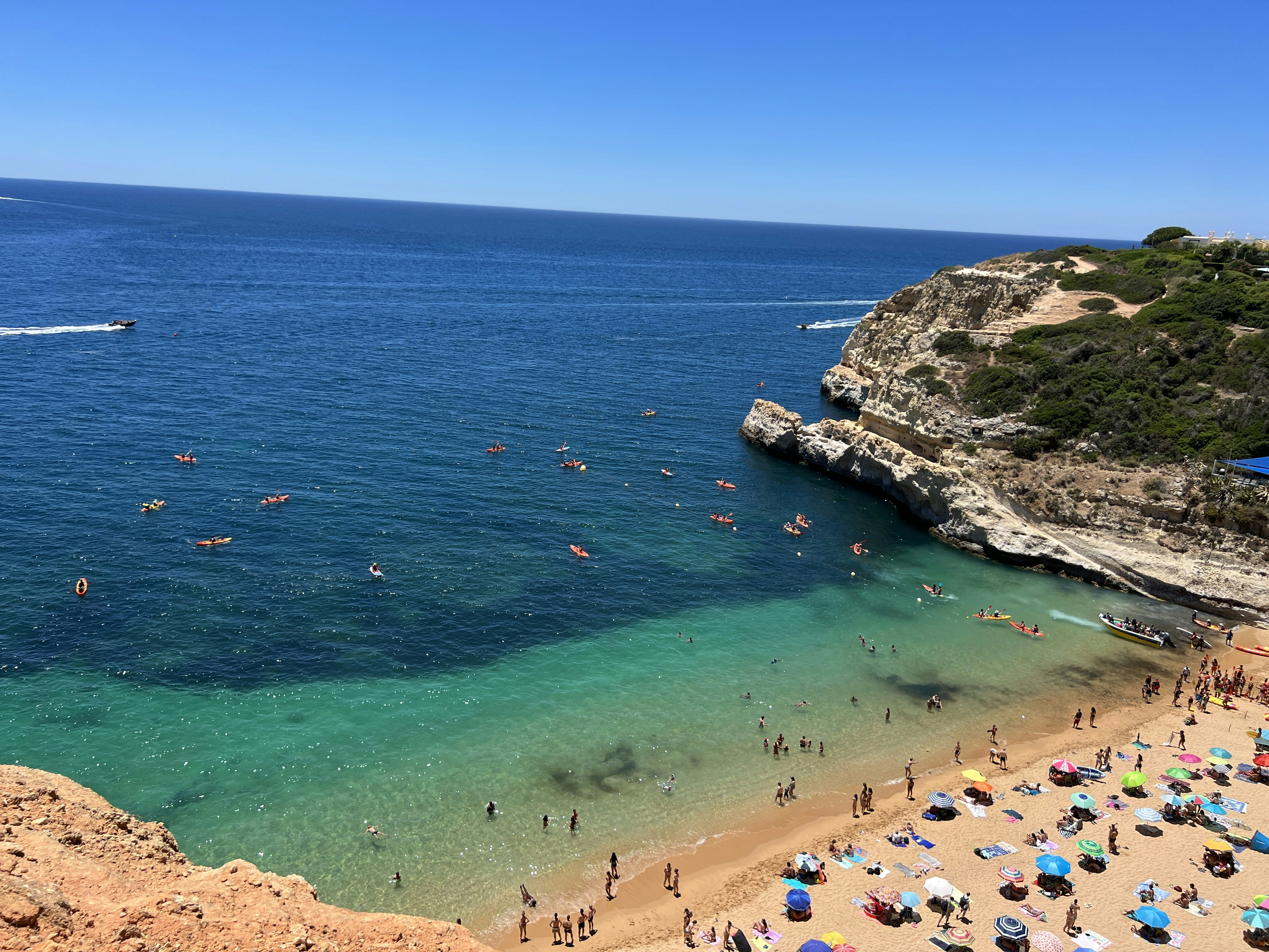 Turquoise waters lap against the golden shore of Benagil Beach in Portugal's Algarve region, as a sailboat glides effortlessly across the calm sea, while sunbathing and swimming enthusiasts soak up the Mediterranean sunshine.