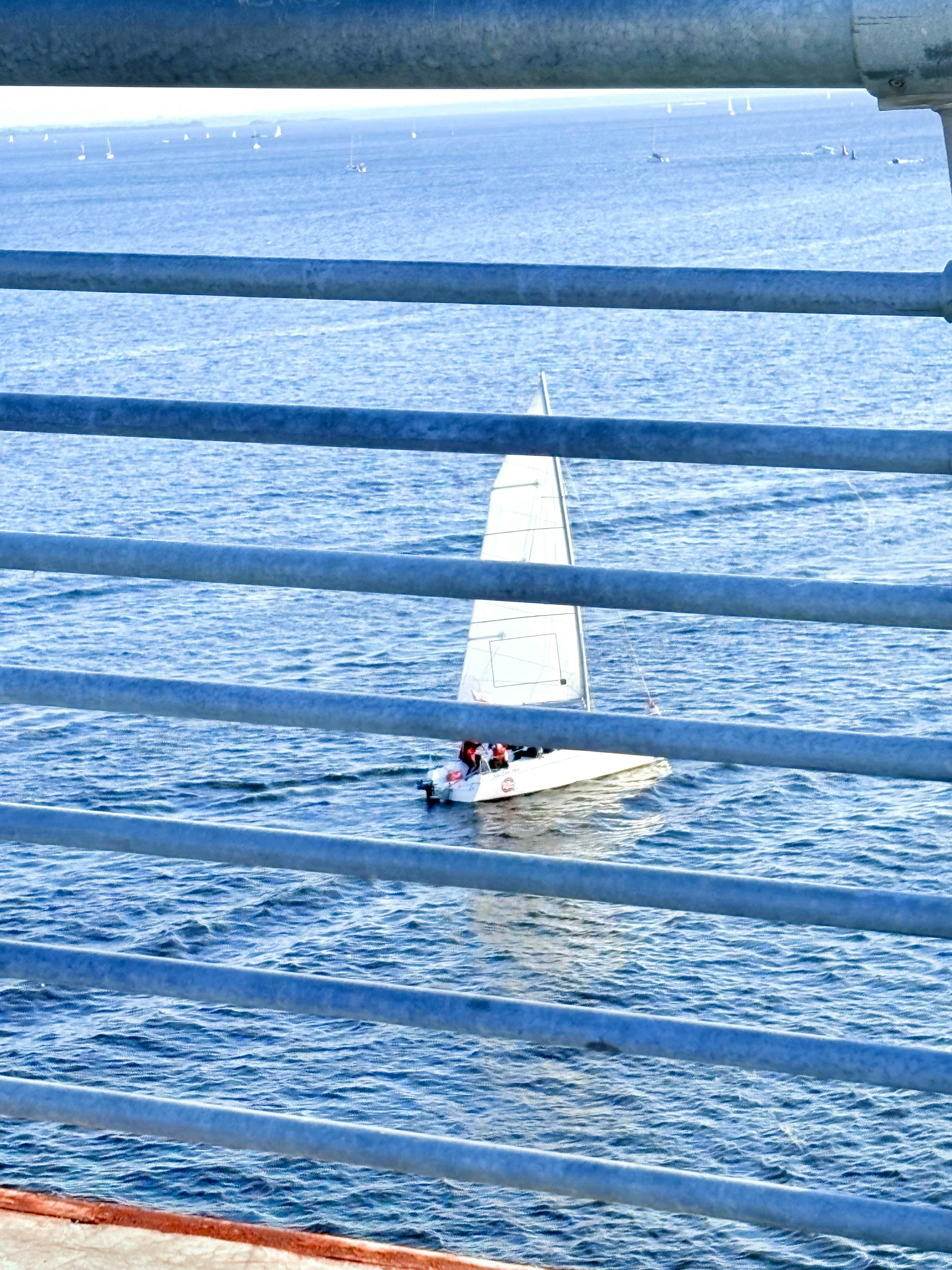 Small sailboat cuts through blue water, viewed through horizontal railing bars; the scene emphasizes open sea and leisure under clear light.