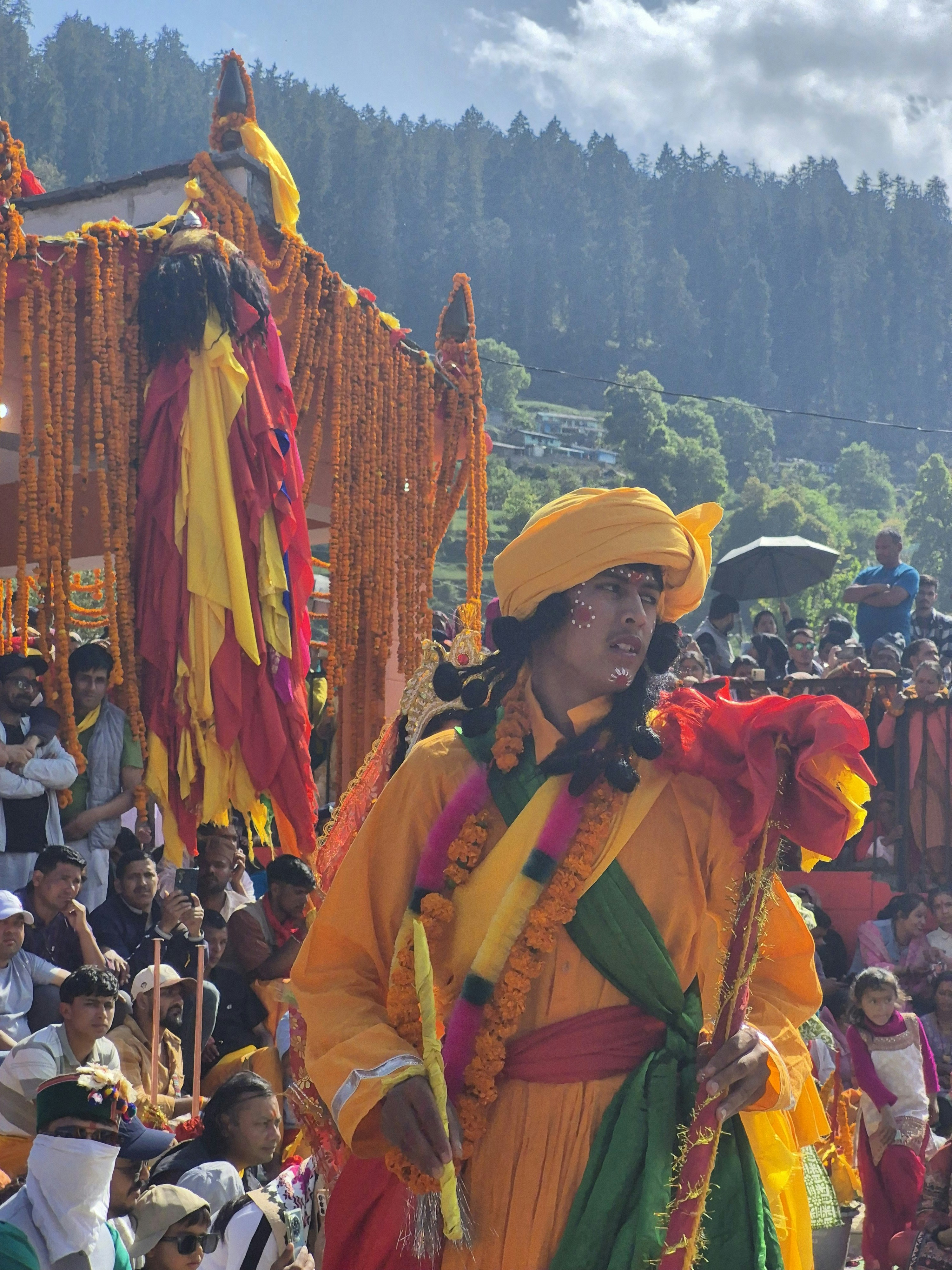 Colorful procession with a central performer in yellow robes, marigold arches, and a crowd in the background; a lively outdoor festival scene.