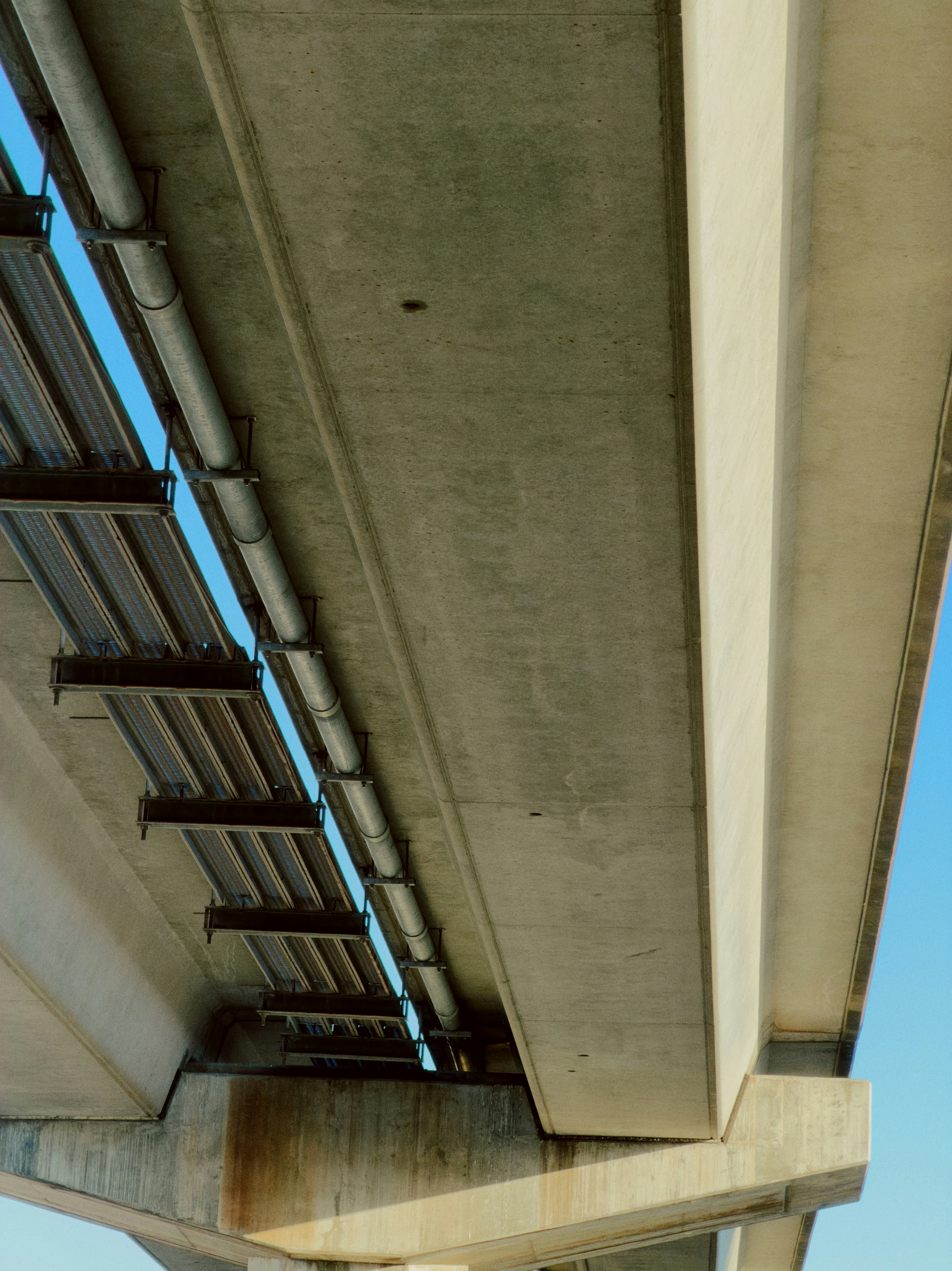 Underside of a concrete bridge with pipes against blue sky.
