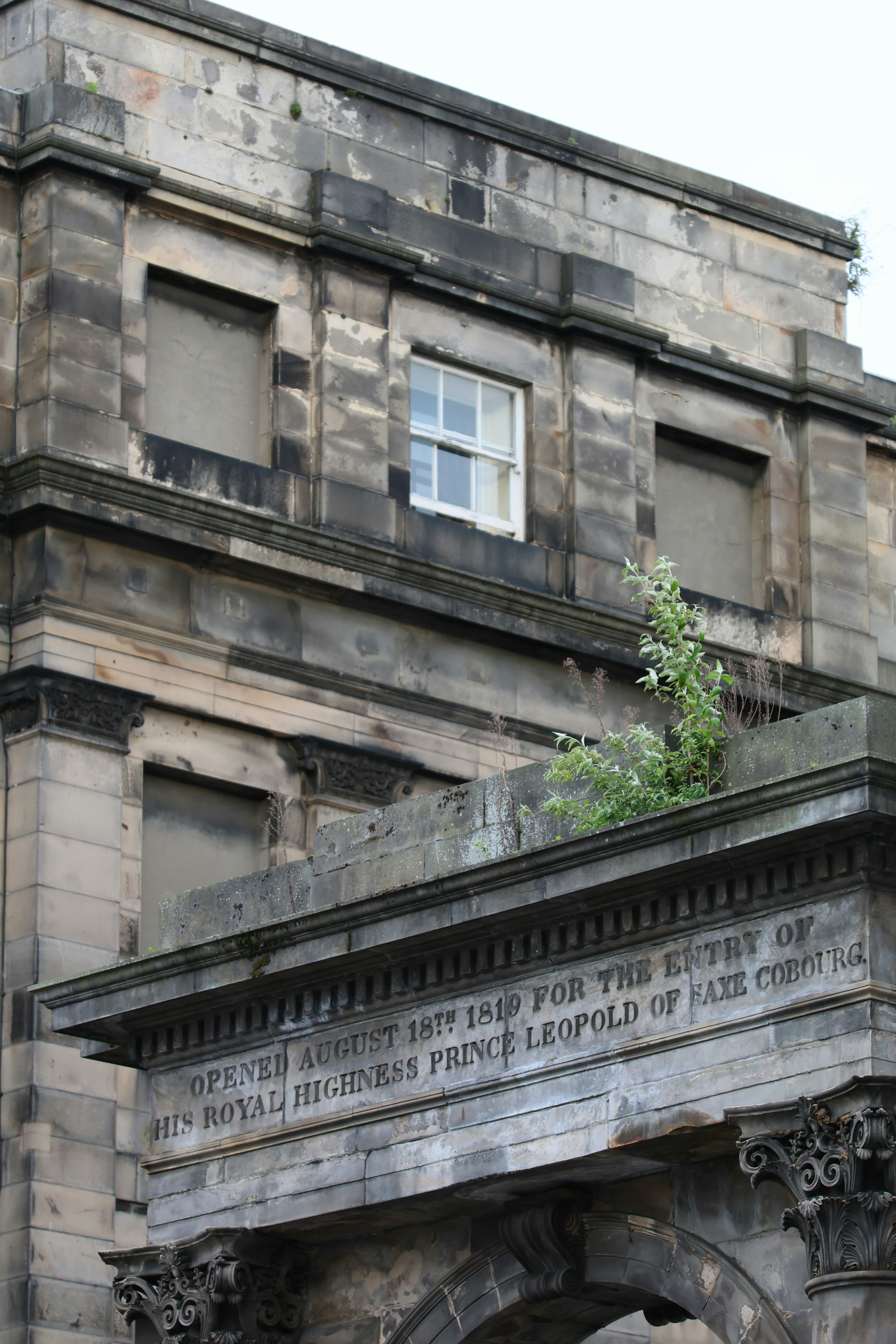 Weathered historic stone building with ornate arch and overgrown greenery on ledge; architectural details highlight erosion and plant life in a neglected urban facade.