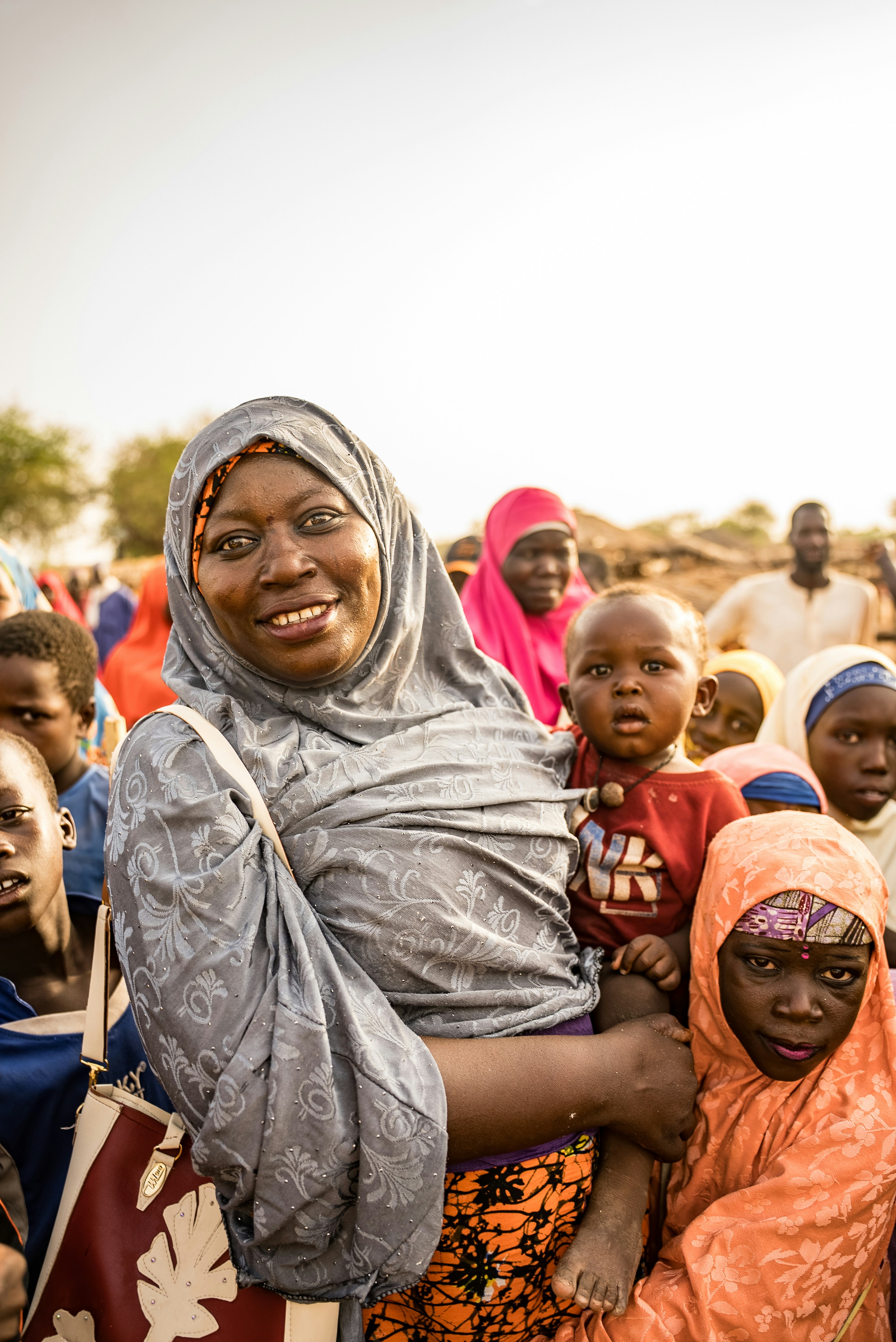 Smiling woman in headscarf holds a child among a crowd.