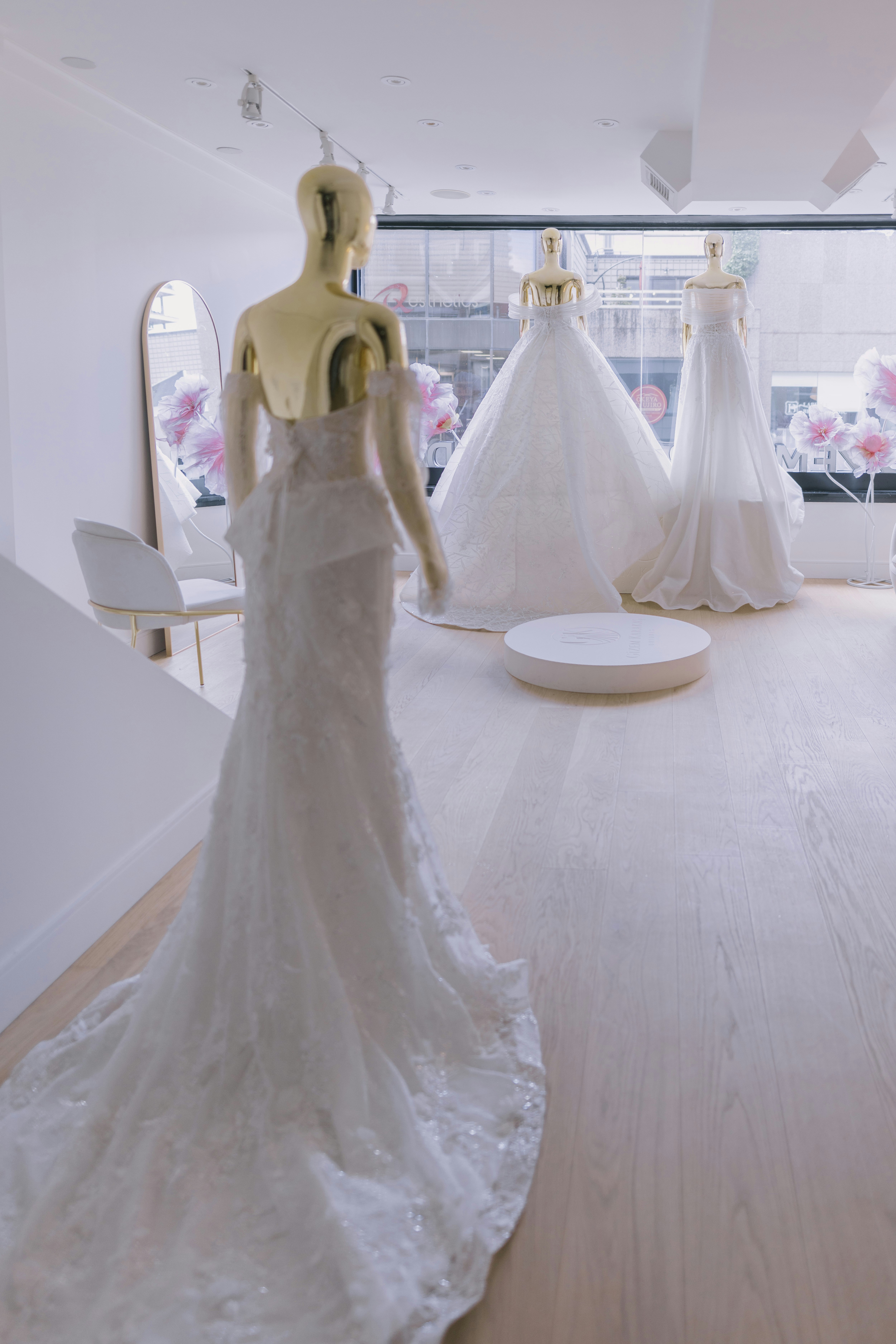 A bright, modern bridal boutique interior featuring stunning white wedding gowns displayed on striking gold mannequins. The natural light from the large front windows illuminates the delicate lace and voluminous tulle skirts against the light hardwood floors.
