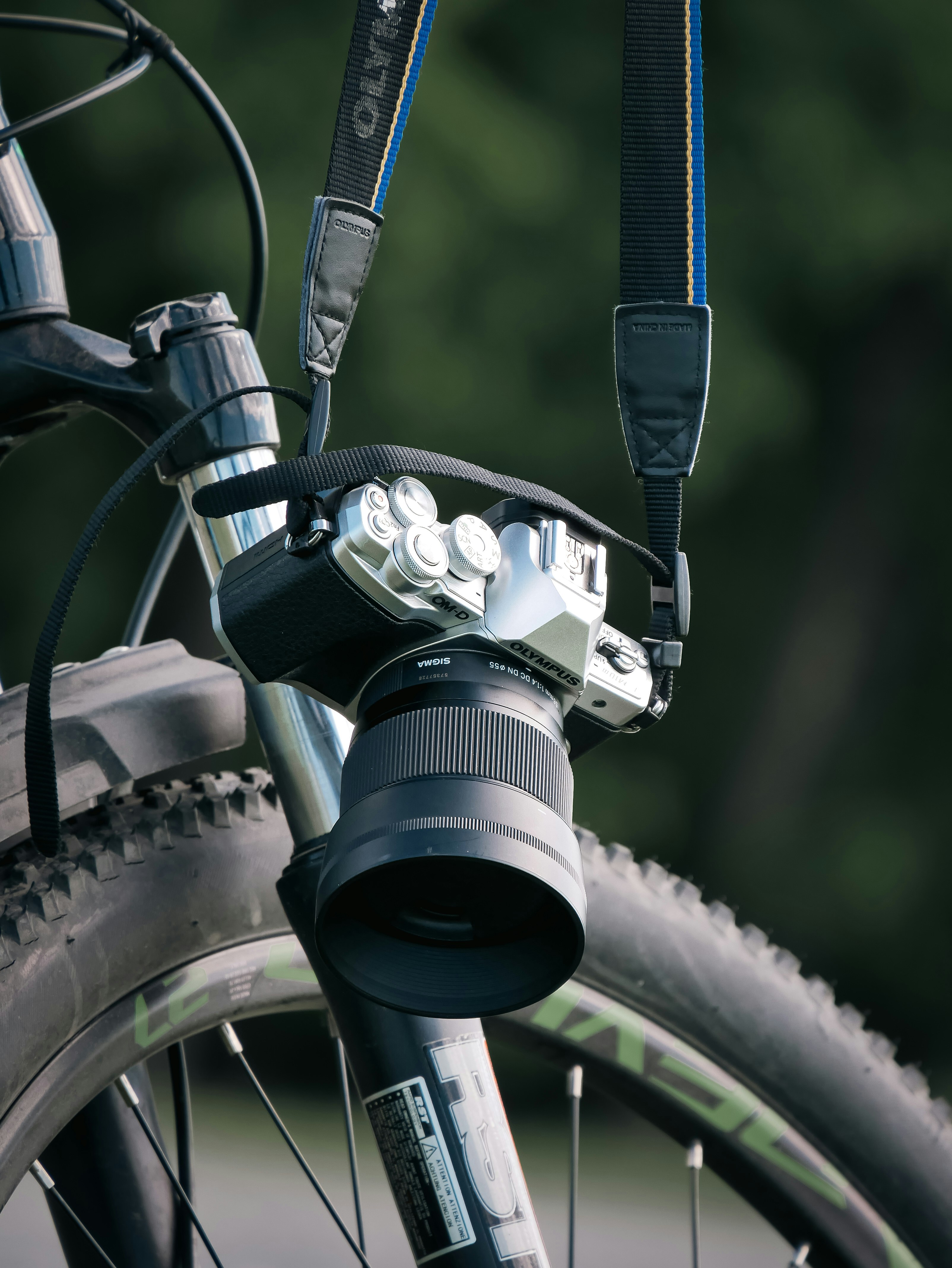 Close-up of a silver camera mounted on a bicycle fork with a large lens, strap and tire details clearly visible.