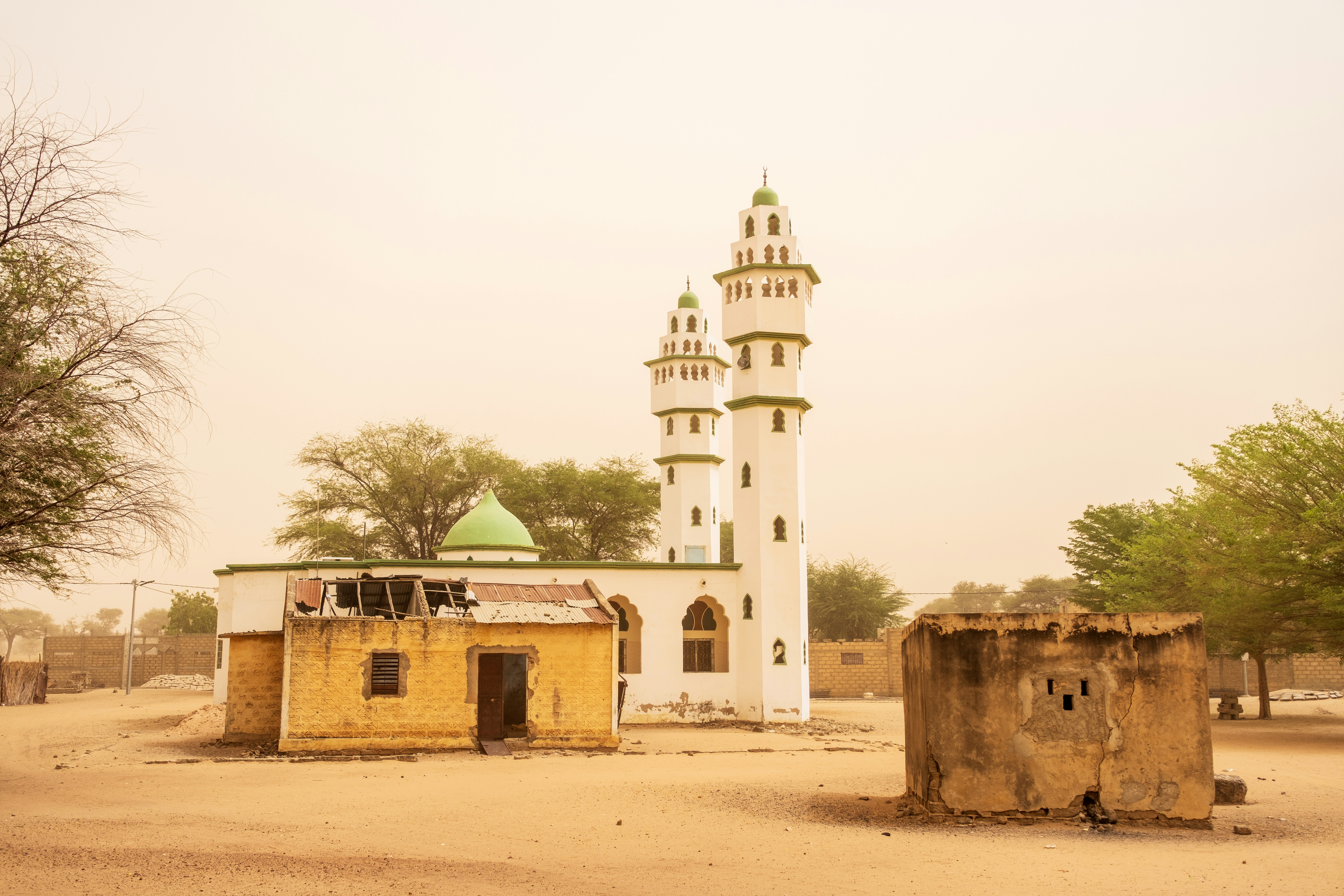 A wide architectural shot of a traditional white mosque with dual minarets and a vibrant green dome, located on the dusty desert road toward Touba, Senegal. The serene structure stands out against an arid, sand-haze sky, juxtaposed with rustic earth-toned foreground buildings and scattered acacia trees typical of the Sahel region.