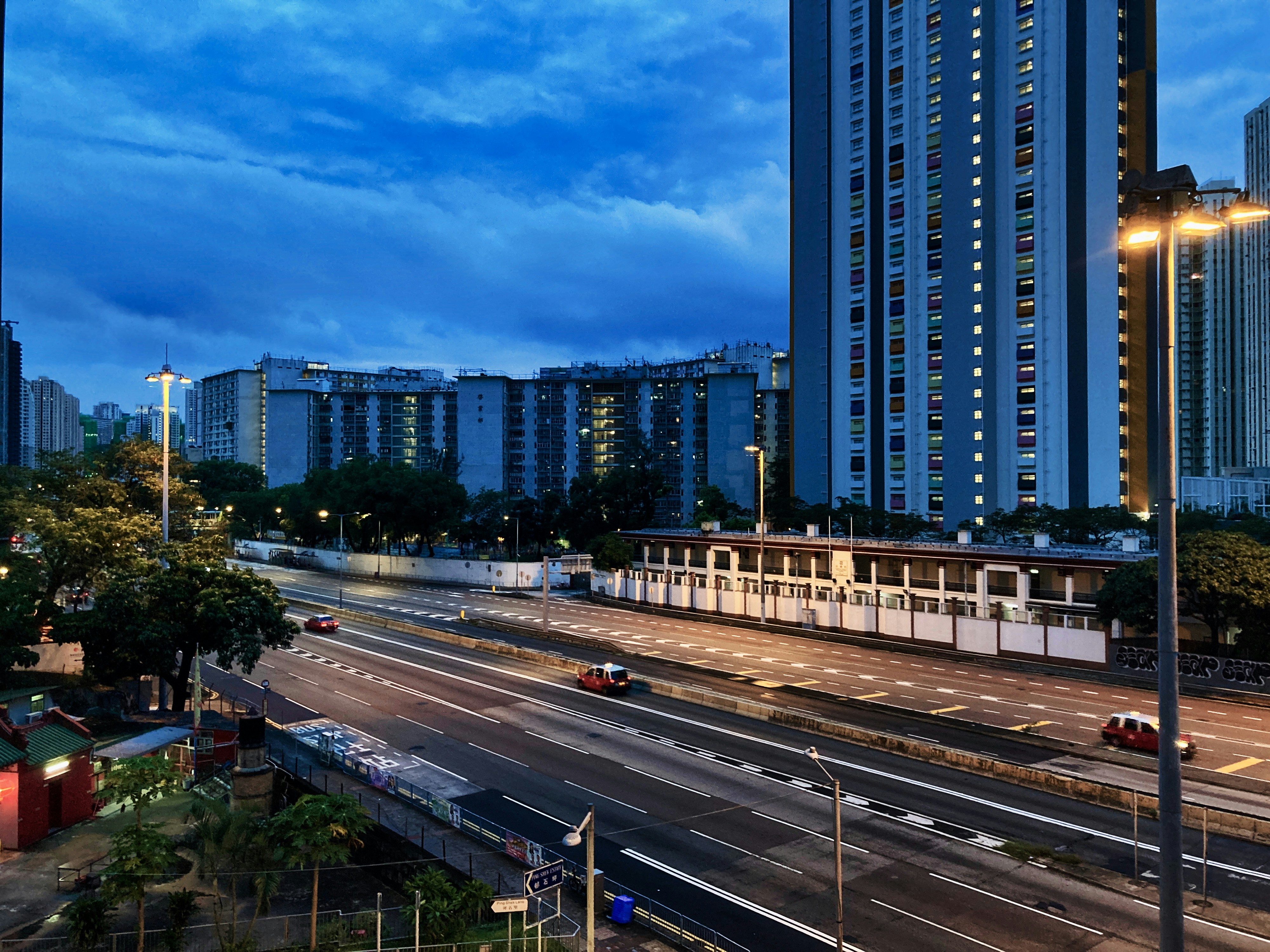 Evening cityscape featuring a multi-lane highway lined with streetlights, flanked by tall residential buildings and a low-rise structure along the roadside.