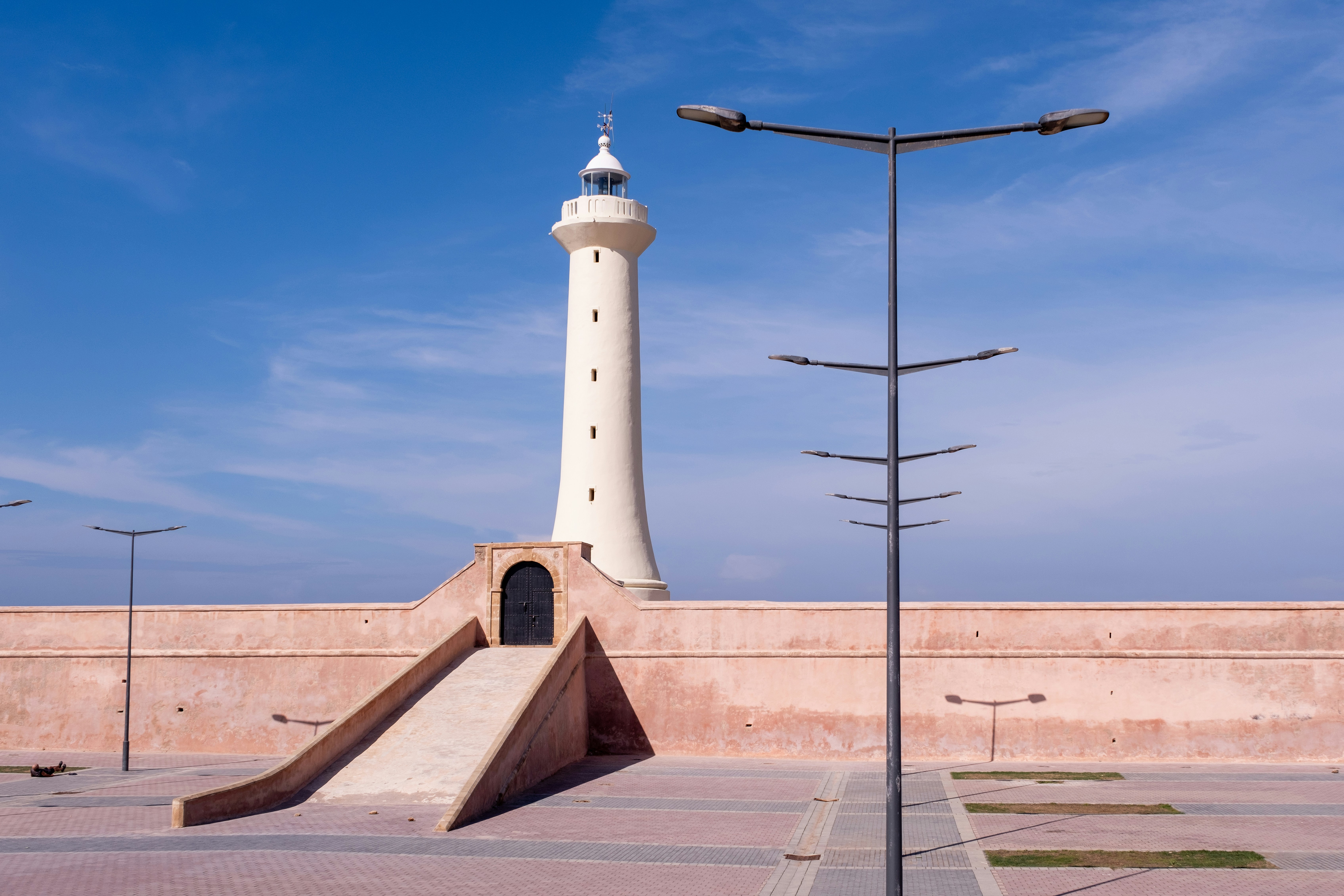 A low-angle, geometric architectural shot of the historic Rabat Lighthouse in Morocco. The tall white stone tower rises behind a warm ocre-toned fortification wall, framed by a concrete ramp, a public plaza pavement, and a modern streetlamp structure against a bright blue sky.