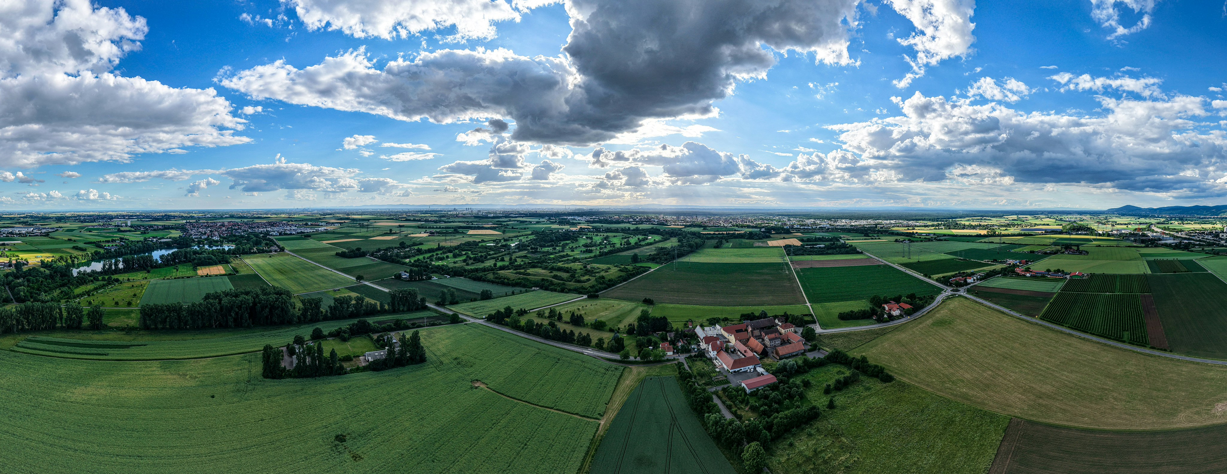Expansive green farmlands with buildings and trees under cloudy sky.